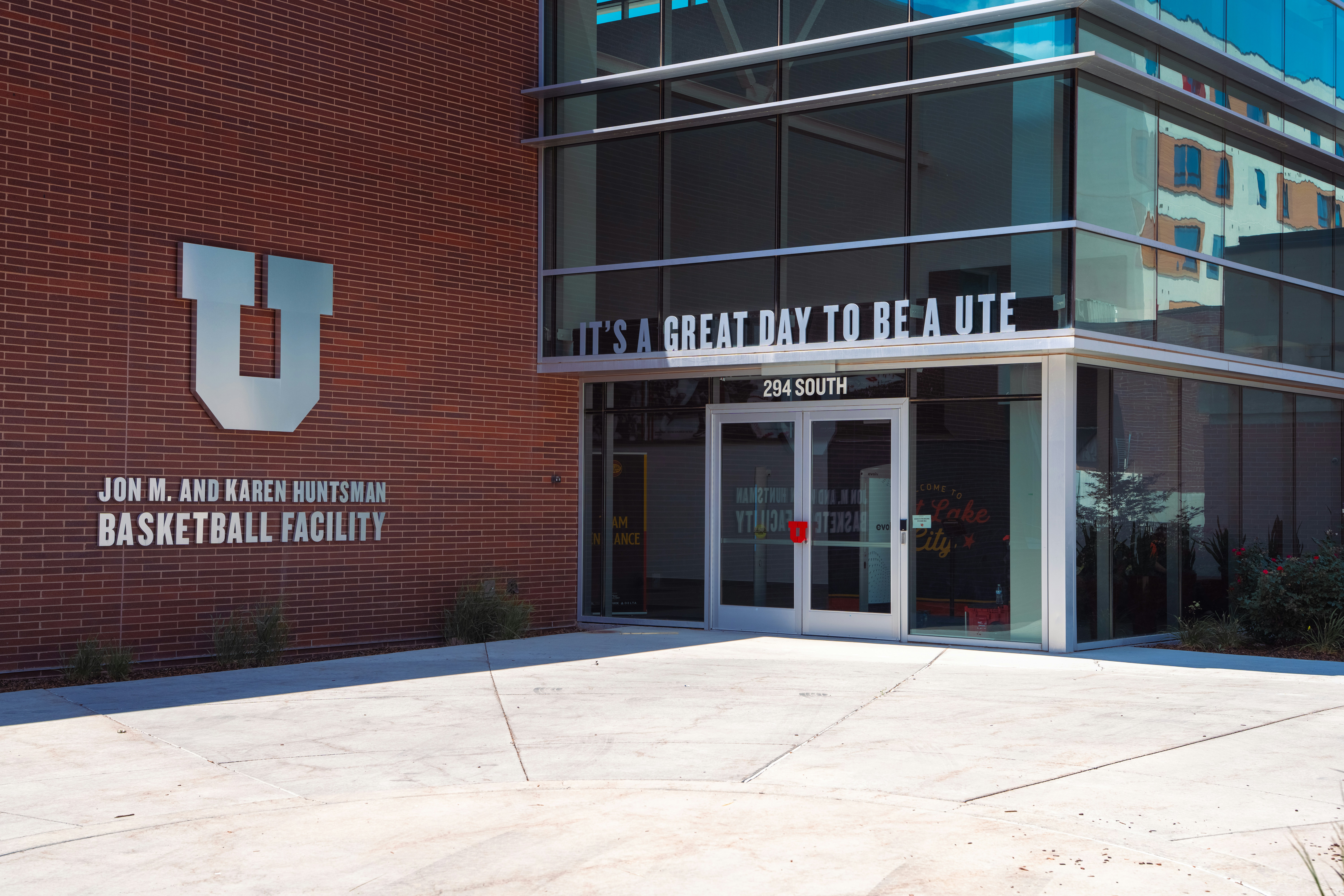 The entrance to the Jon M. and Karen Huntsman Basketball Facility at the University of Utah features a bold red-brick exterior, large glass windows, and the university’s block “U” logo. Above the doors, the slogan “It’s a great day to be a Ute” is prominently displayed, welcoming visitors and athletes. | Modern building entrance with large u logo