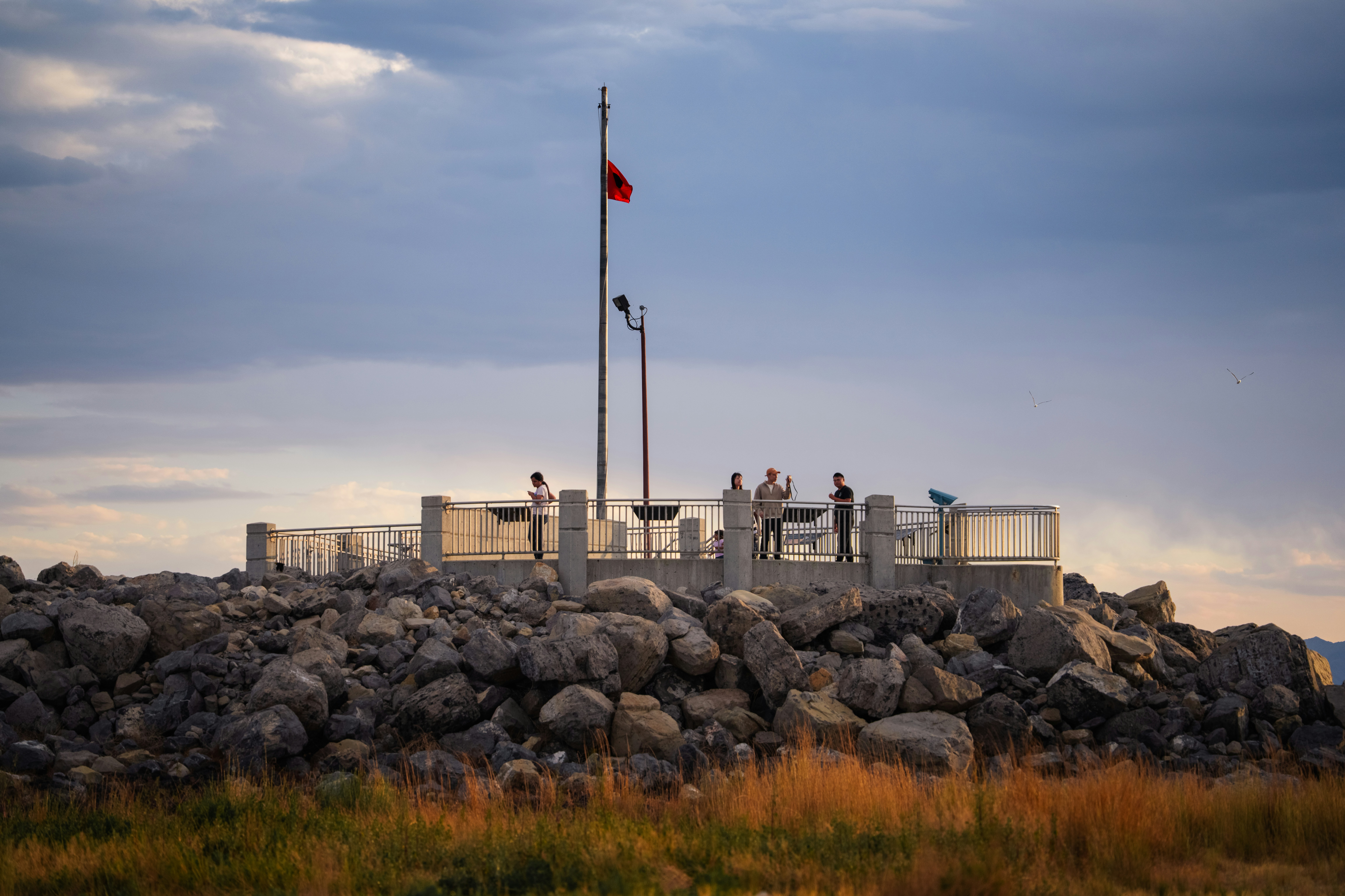 A small group of people stands on a concrete observation platform surrounded by large boulders at the edge of the Great Salt Lake. A red flag flutters at half-mast under a cloudy evening sky, with golden light touching the grasses in the foreground. | Flagpole on a rocky hill at dusk