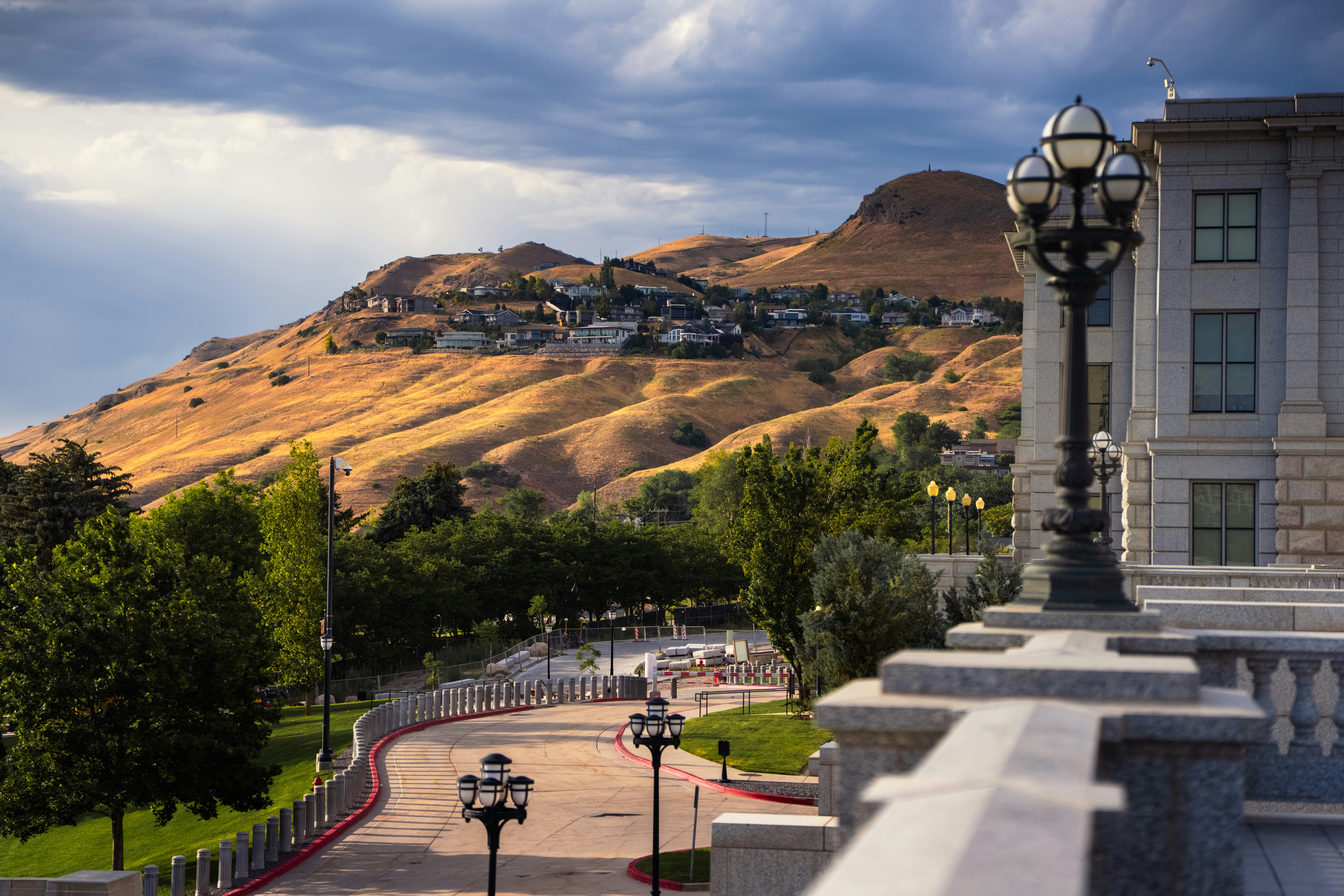A scenic view showcasing a hillside community framed by lush trees and a winding pathway, with dramatic clouds casting shadows over the landscape.