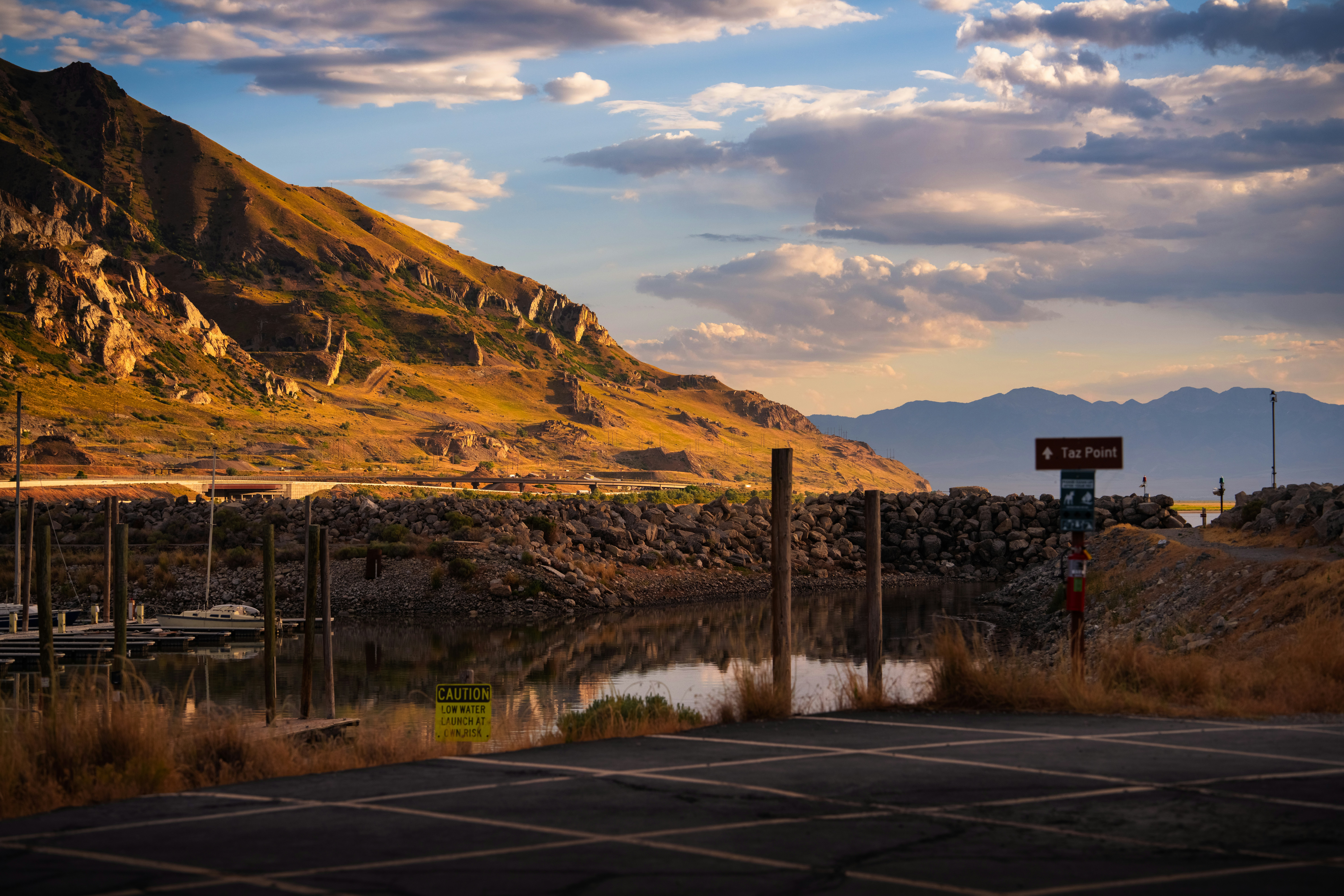 A tranquil lakeside scene featuring a mountain backdrop and a wooden dock. The sun casts a warm glow on the landscape, highlighting the natural beauty of the area.