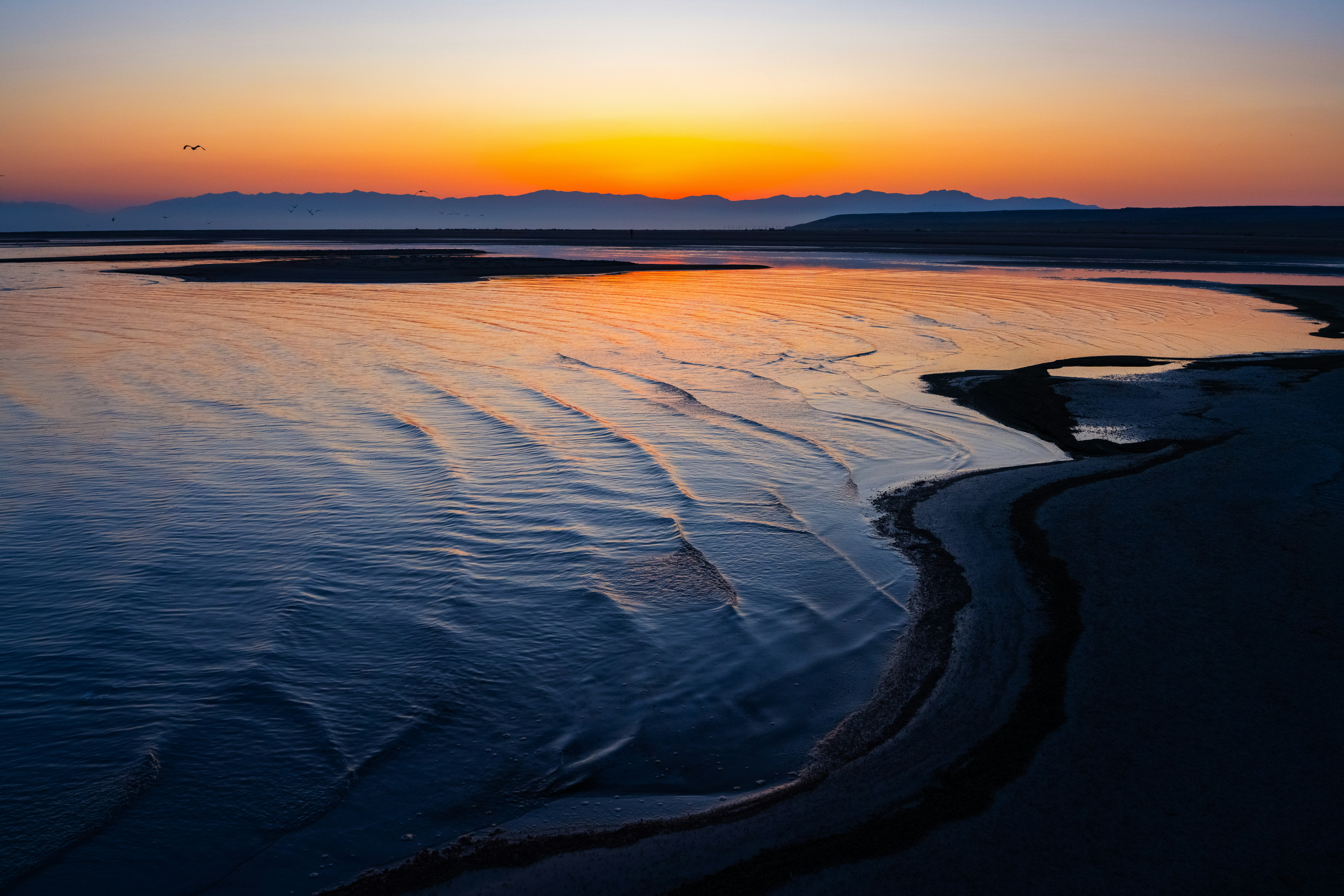 A tranquil shoreline along the Great Salt Lake at sunset glows with vibrant orange and deep blue hues, the fading light reflecting softly across gentle ripples in the water. The curving sandbar in the foreground adds a sense of depth, while distant mountains form a dark silhouette against the fiery Utah horizon. | Sunset over a calm ocean