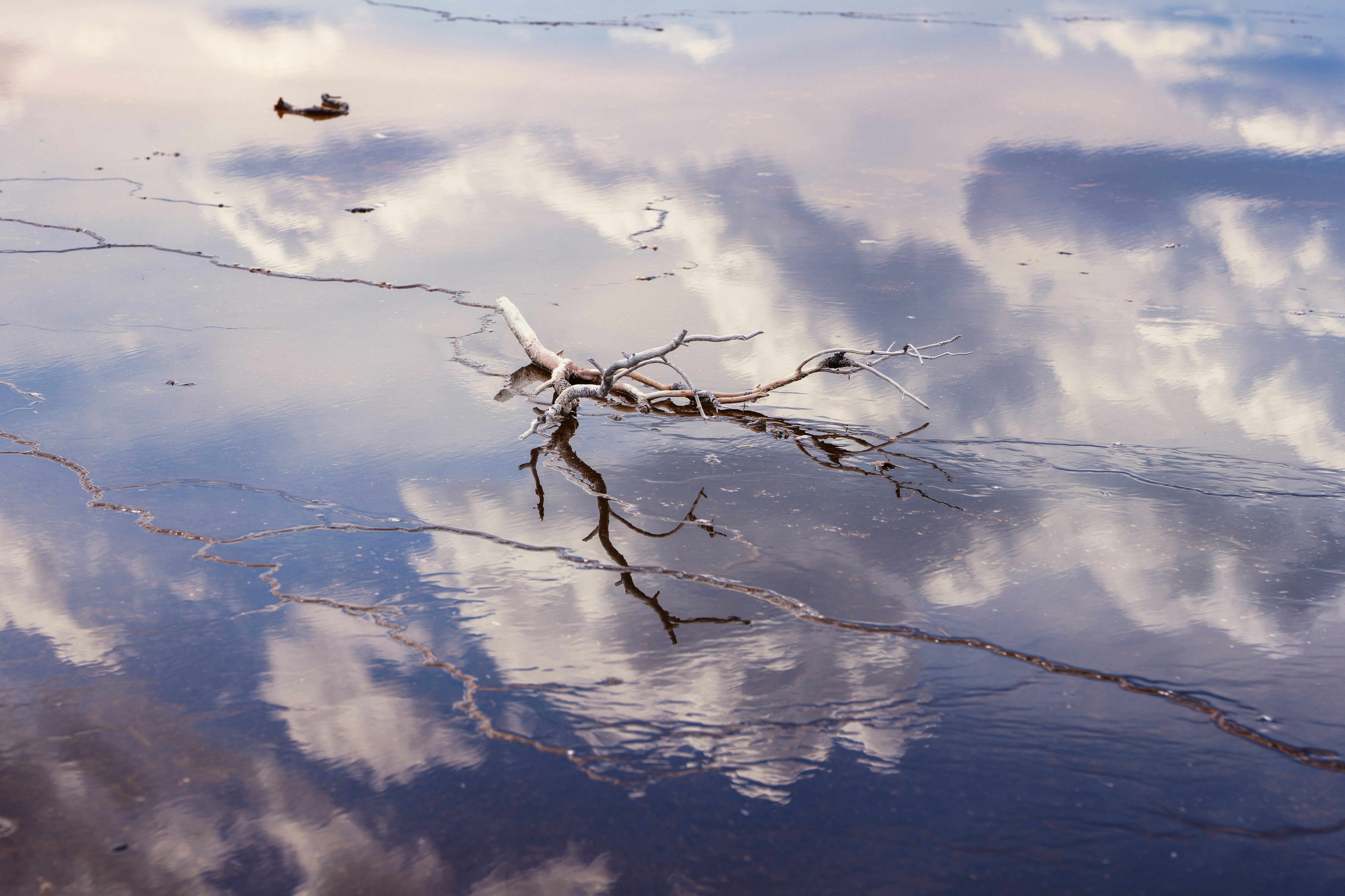 A lone branch rests on the reflective surface of a hot spring in Yellowstone National Park, mirroring the sky’s fluffy white clouds and soft blue tones. Subtle mineral lines ripple across the water, adding delicate texture to the serene scene. | Airplane flying through dramatic clouds over mountains
