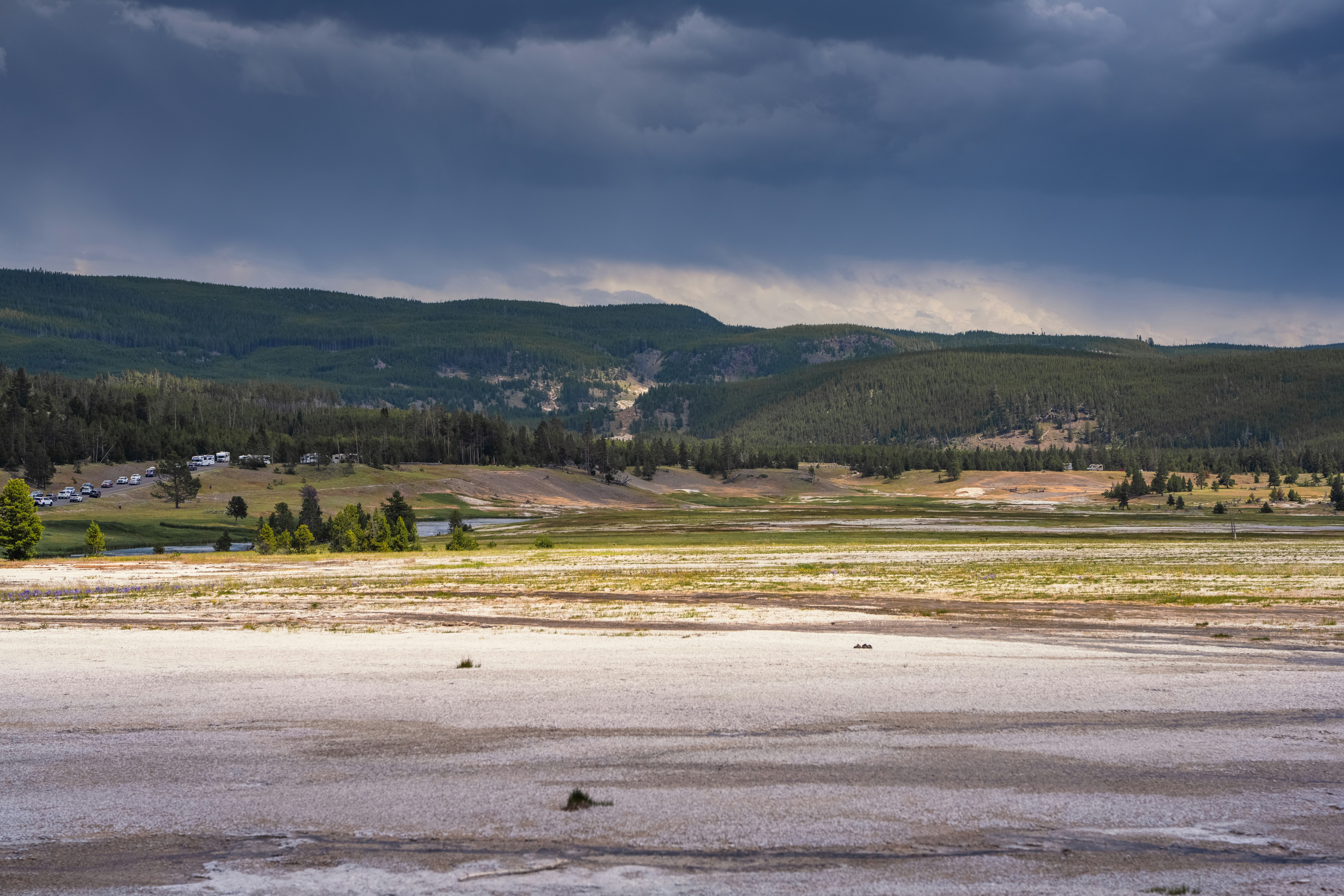 A wide view of Yellowstone National Park’s geothermal basin under a moody, stormy sky, with pine-covered hills and parked vehicles in the distance.