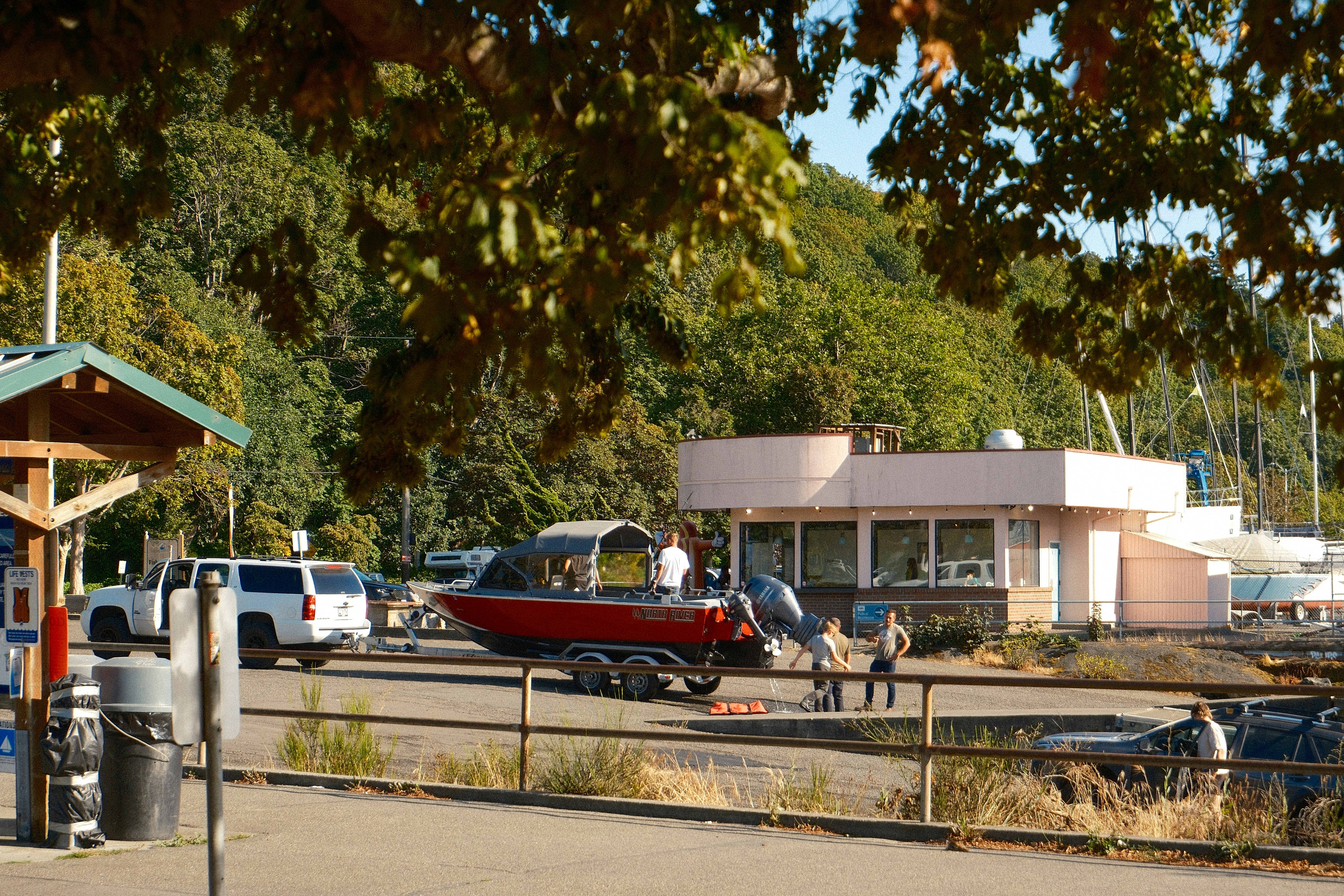 Boat being prepared for launch at a bustling marina, surrounded by lush greenery and a pastel-colored building. 