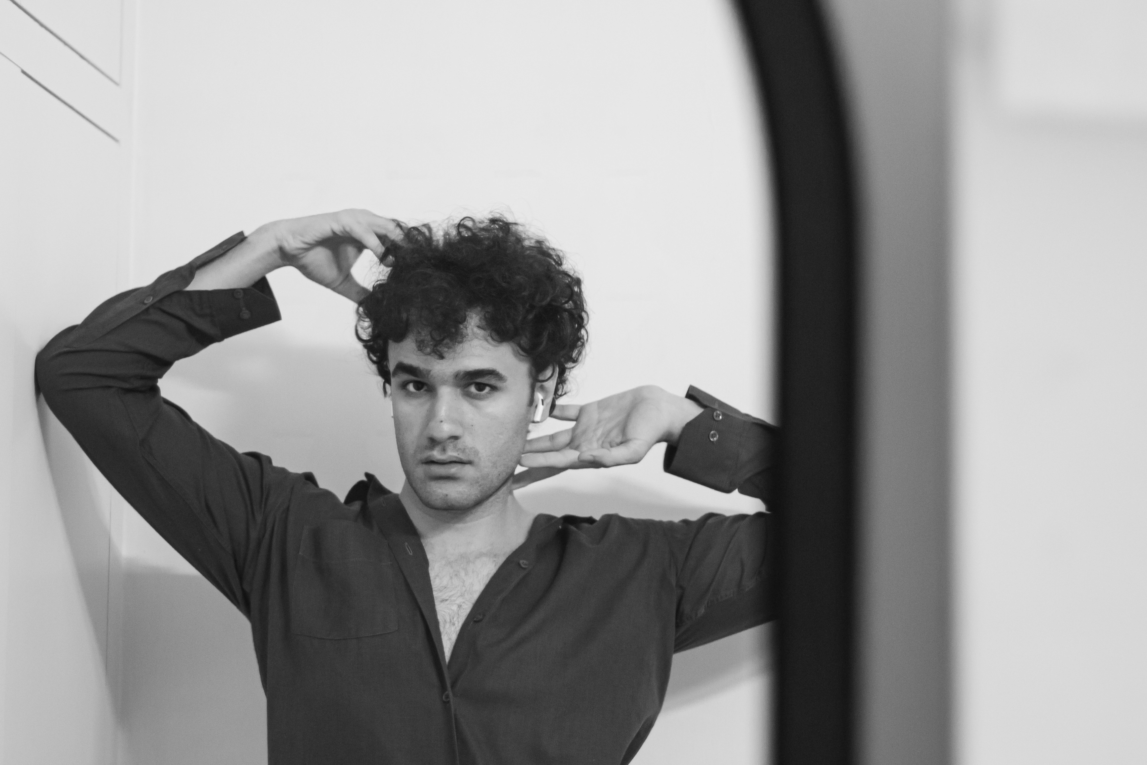 A young man poses thoughtfully in a minimalist setting, framed by a mirror that adds depth to the composition.