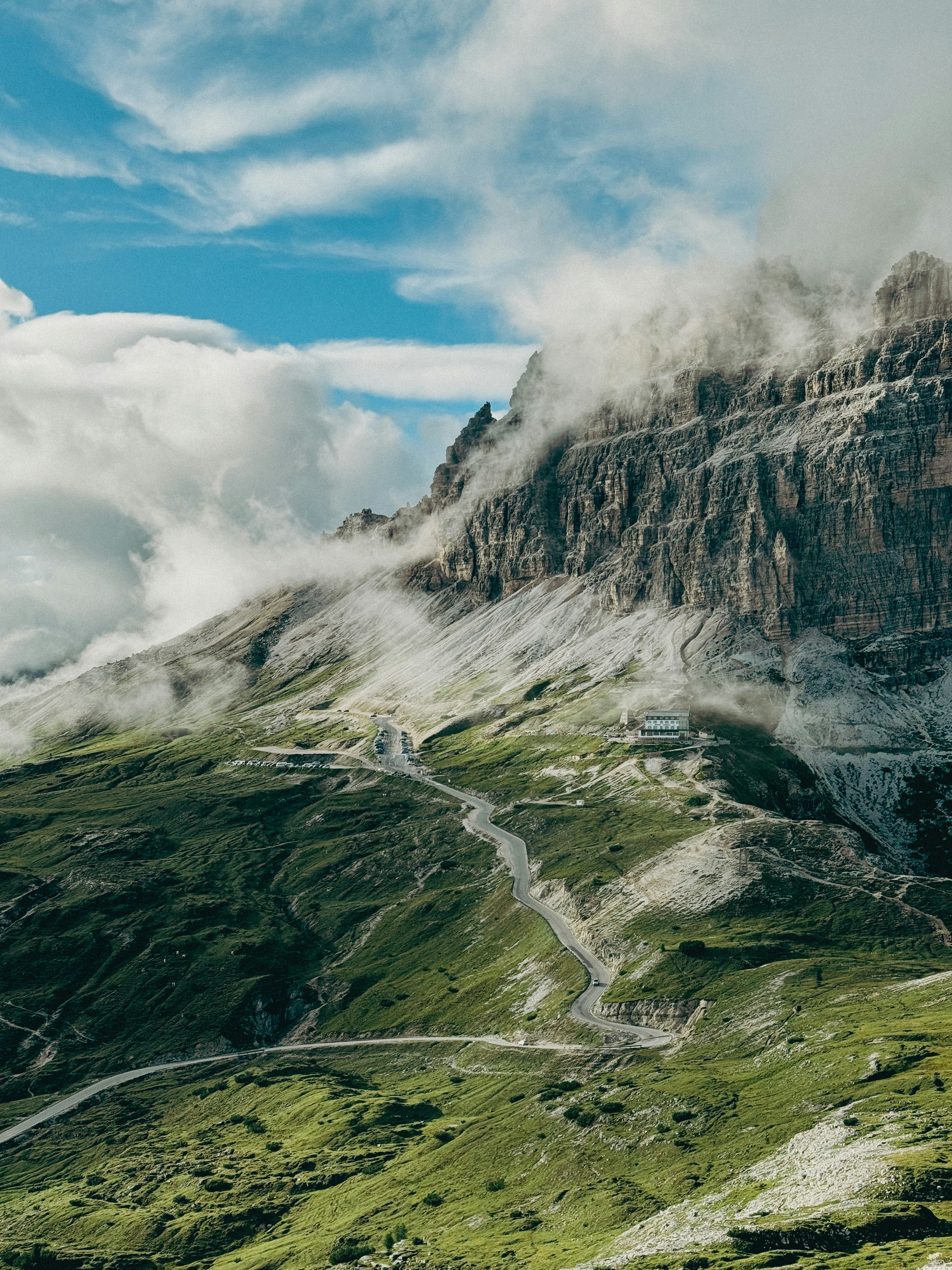 Majestic mountain landscape with a winding road and a distant building, enveloped in clouds and greenery. Perfectly captures the serene beauty of the Dolomites.