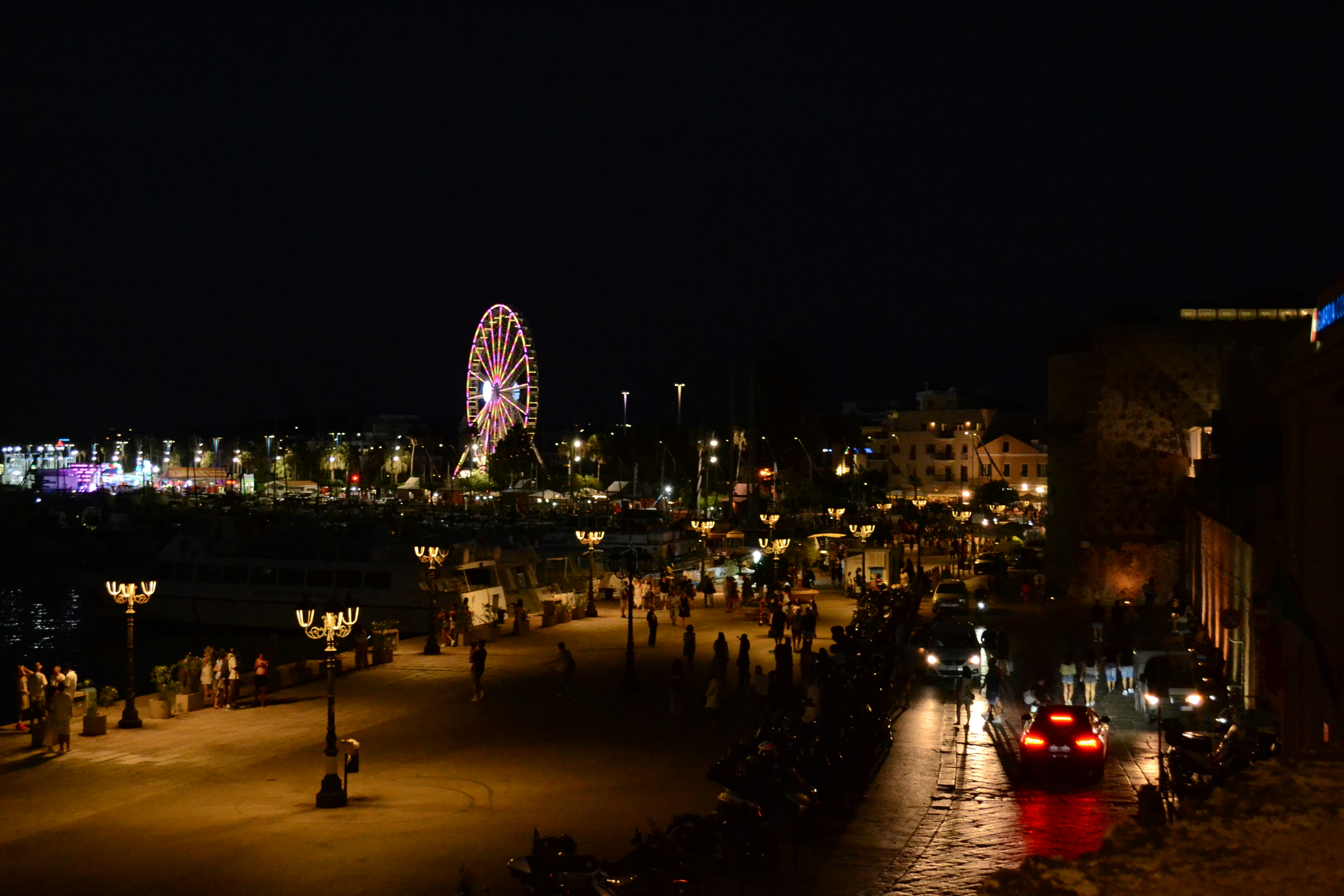 A vibrant waterfront scene at night featuring a colorful Ferris wheel, bustling pedestrians, and shimmering reflections on the water.