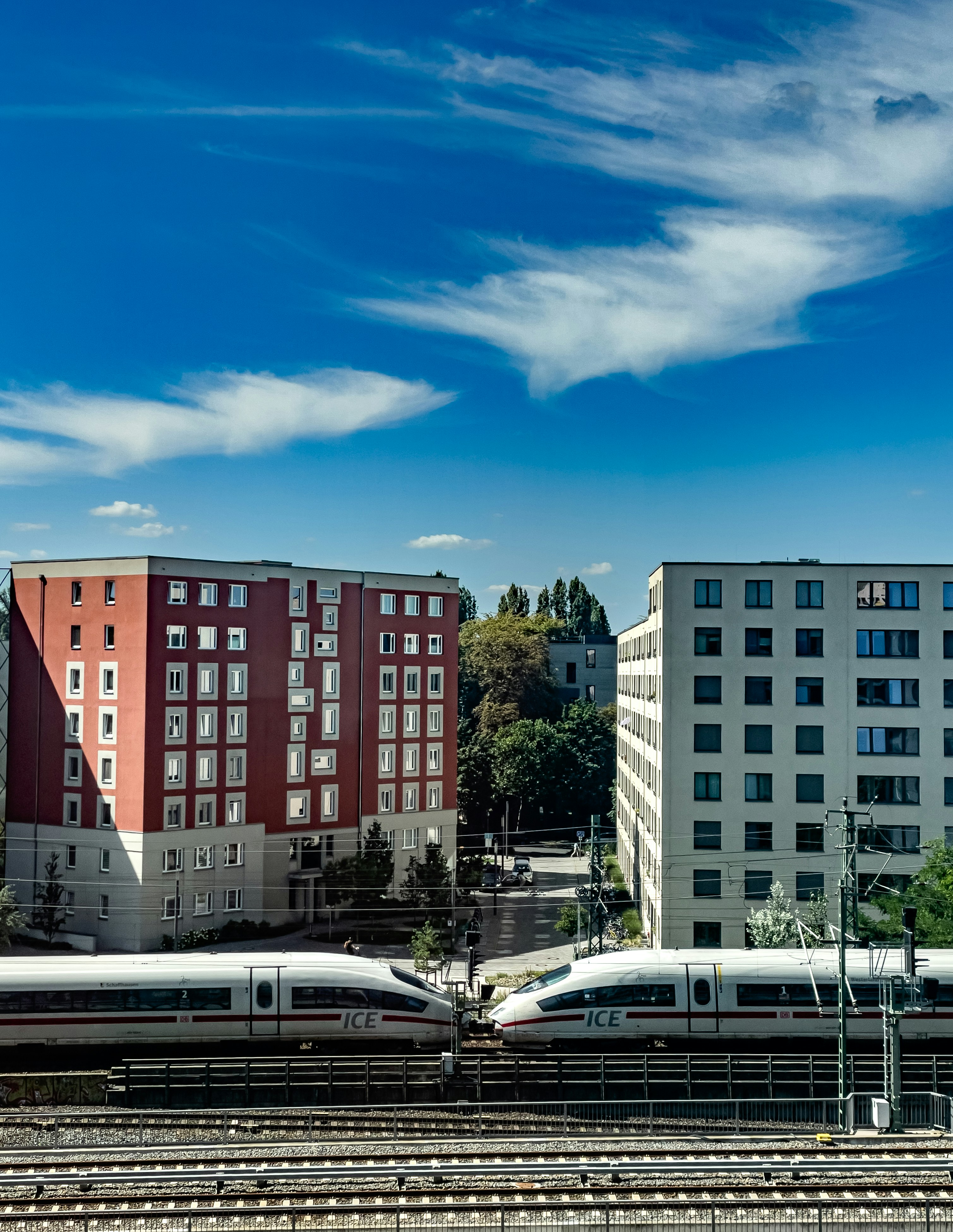 Two modern trains pass by apartment buildings under a blue sky.