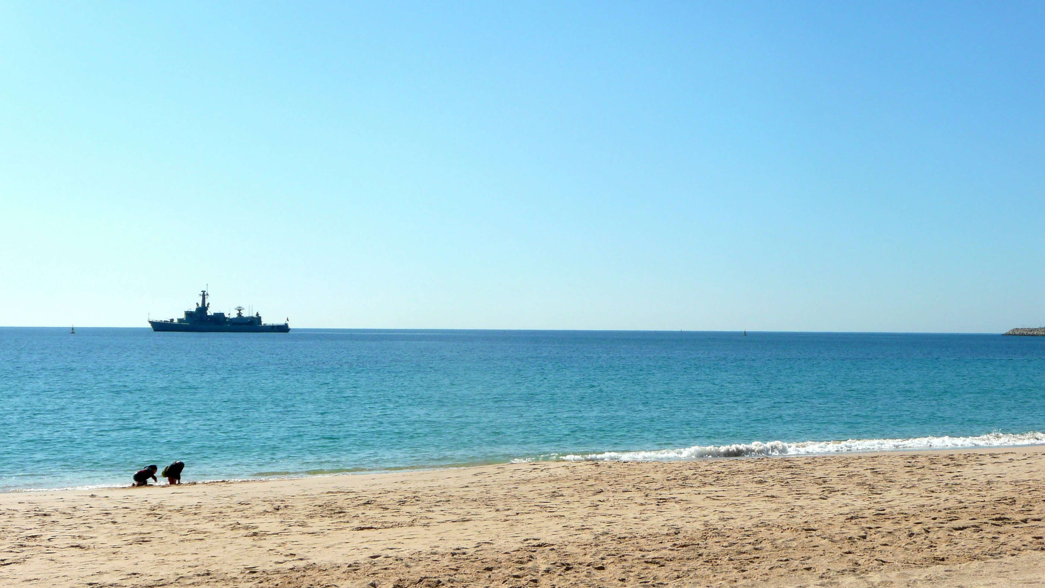 On a quiet stretch of golden sand, two figures crouch at the water’s edge, lost in the simple joy of discovery. Beyond them, the vast Atlantic glistens under a flawless sky, its calm surface interrupted only by the steady silhouette of a naval ship. A moment where innocence meets duty, and leisure coexists with vigilance — all framed by the timeless rhythm of sea and shore. | A boat sails on the calm blue ocean near shore.
