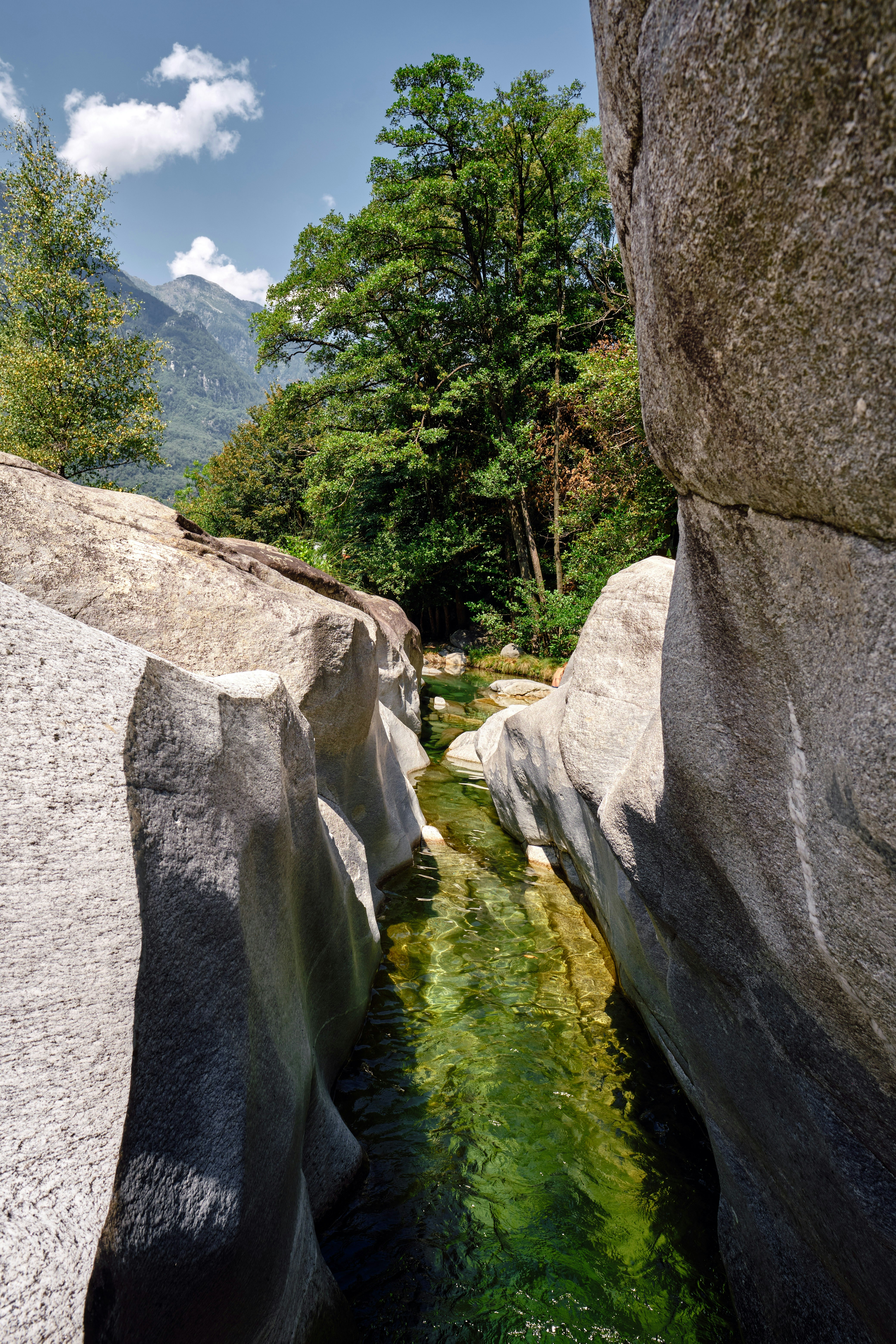 Clear stream winding through smooth rock formations under a bright blue sky, framed by lush greenery.
