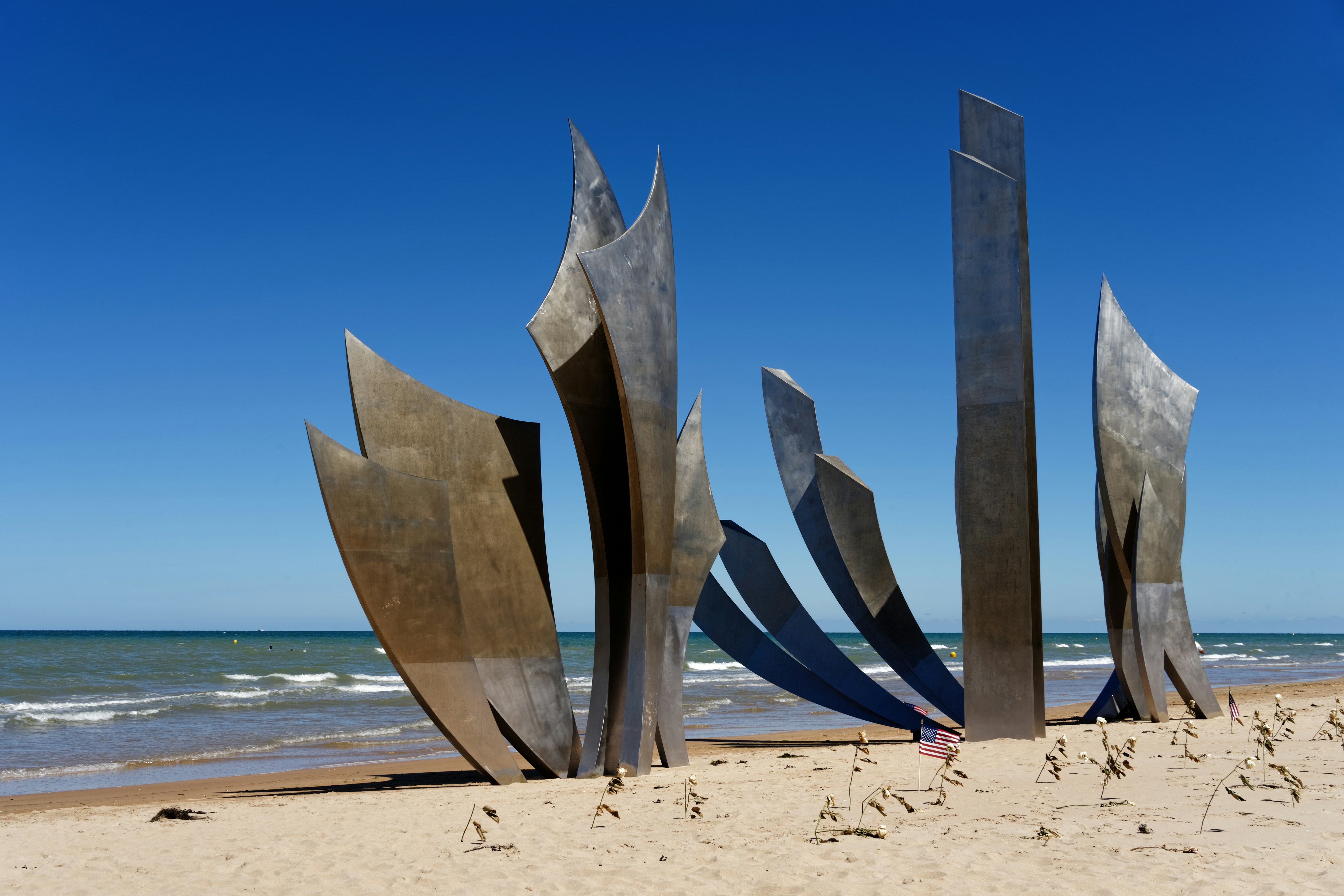 Abstract metallic sculpture rising from sandy beach, casting dynamic shadows against the vibrant blue sky and ocean. 