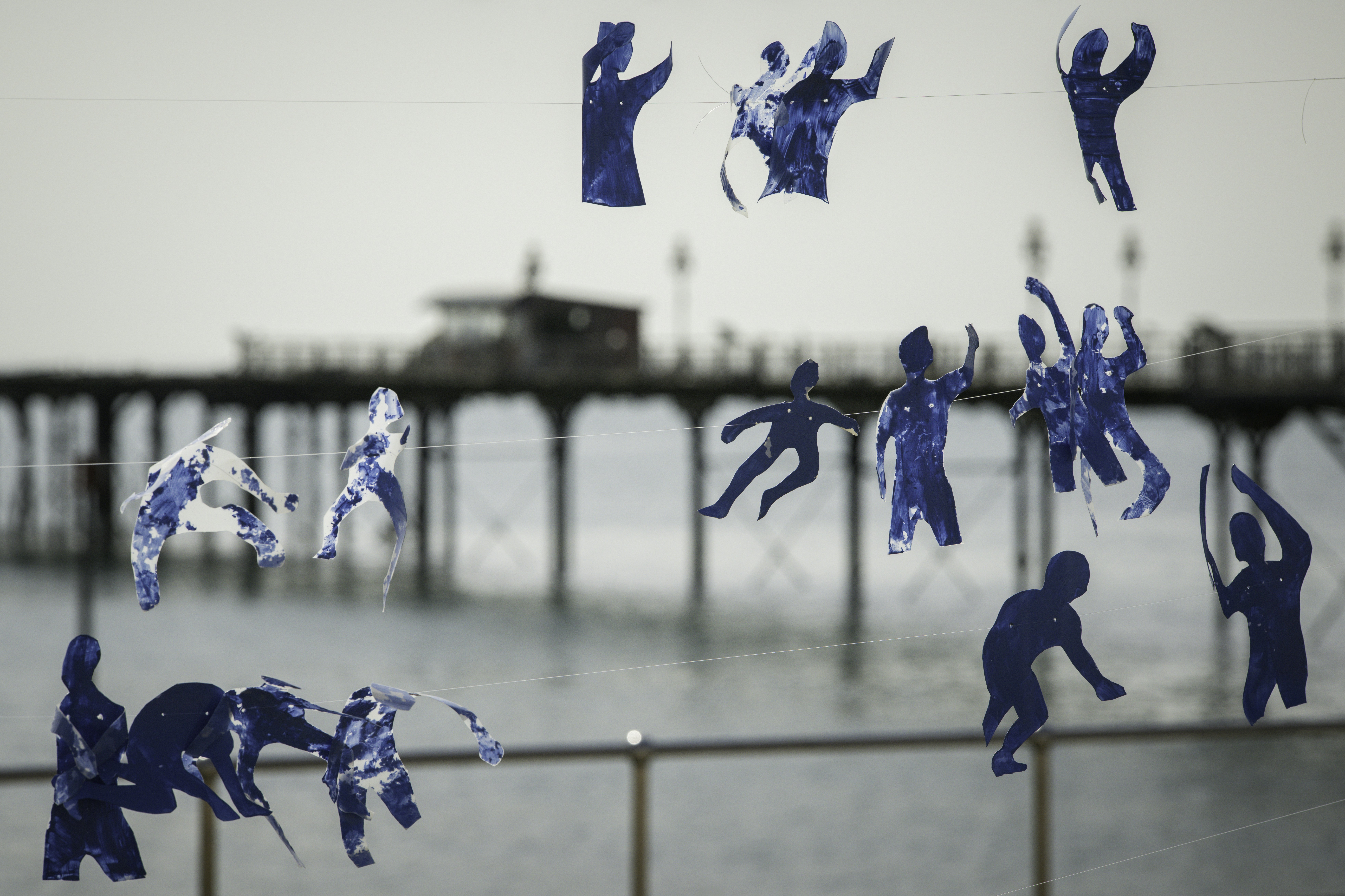 Blue cut-out figures suspended in mid-air, creating a dynamic scene against a coastal pier. The artwork captures movement and playfulness.