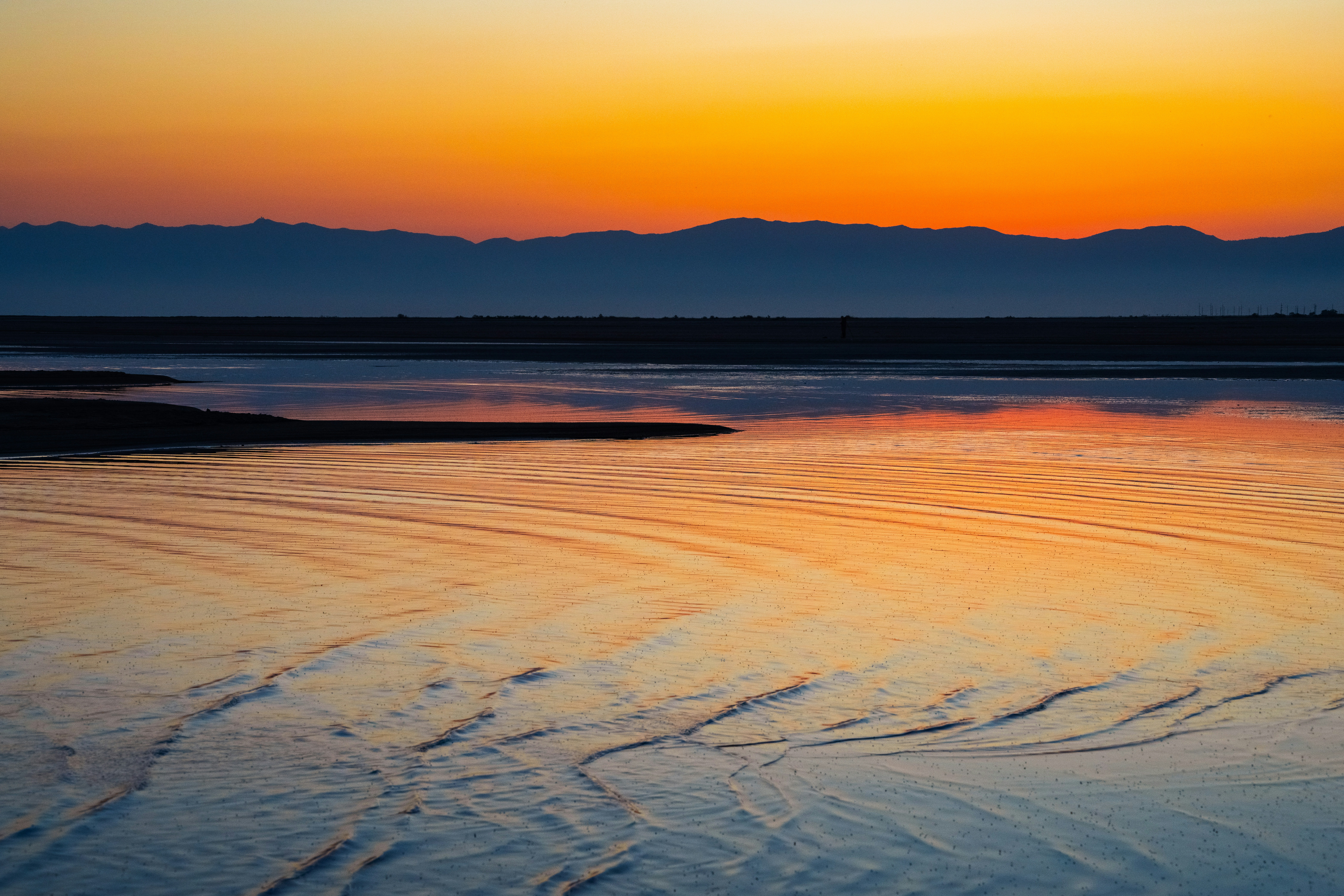 A tranquil lakeshore scene at sunset, where soft ripples on the water reflect a brilliant gradient of colors from golden orange to deep blue. The distant silhouette of mountains contrasts against the glowing horizon, and the stillness of the water enhances the peaceful, meditative atmosphere. The shoreline’s dark shape adds depth to the composition. | Sunset over calm water with distant mountains