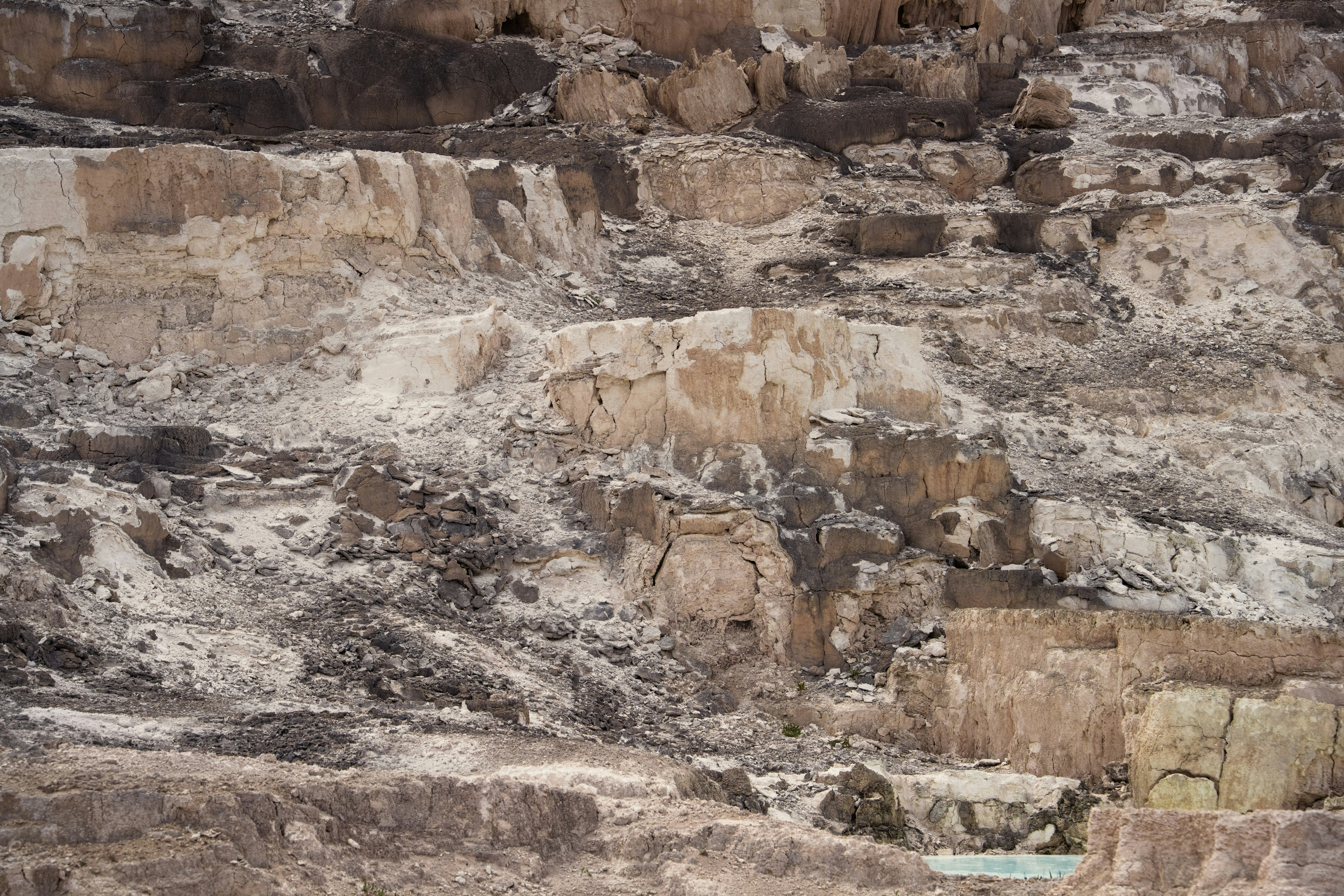 A close-up view of eroded travertine rock formations at Mammoth Hot Springs in Yellowstone National Park. The scene features layers of pale beige, cream, and dark brown stone, shaped by years of geothermal activity and mineral deposition. The rugged texture and fractured surfaces reveal the geological history of the area, with a glimpse of turquoise thermal water at the bottom edge. | Eroded rock formations with layered textures and colors