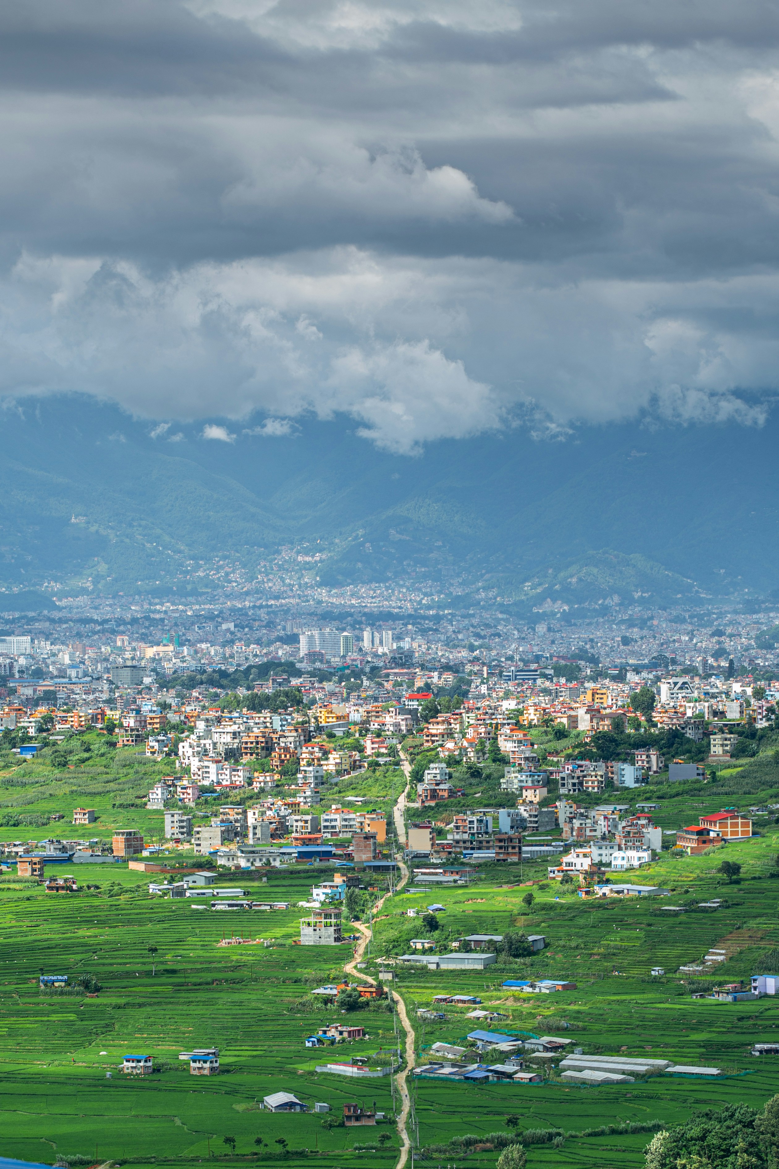 Urban Expansion Meets Verdant Farmlands Under Moody Skies | Cityscape nestled in rolling green hills under cloudy sky
