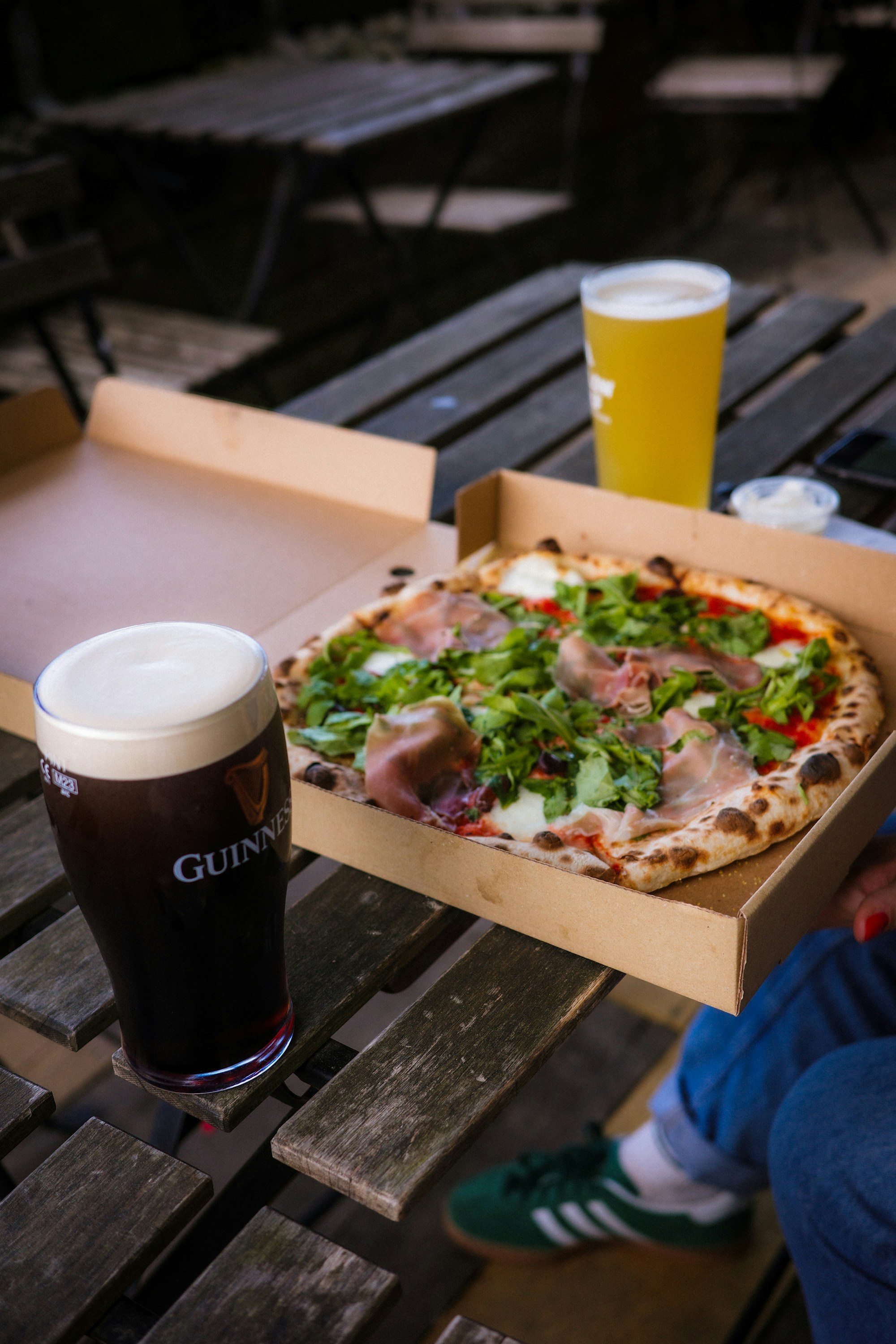 Freshly made pizza topped with greens and cured meats, accompanied by a dark stout and a pale ale, set on a rustic wooden table.
