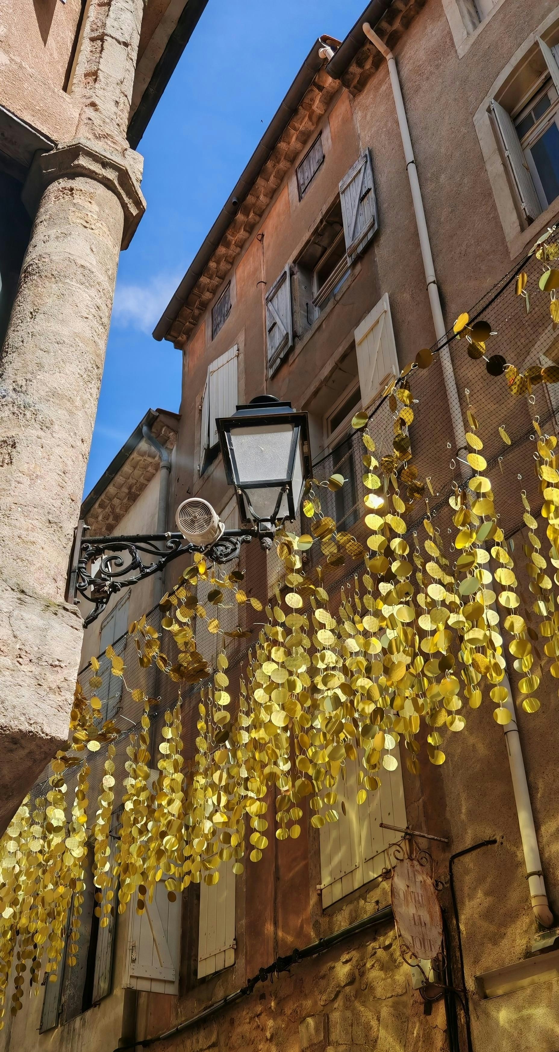 Golden leaves cascade from a lantern in a quaint alleyway, framed by rustic buildings under a clear blue sky.