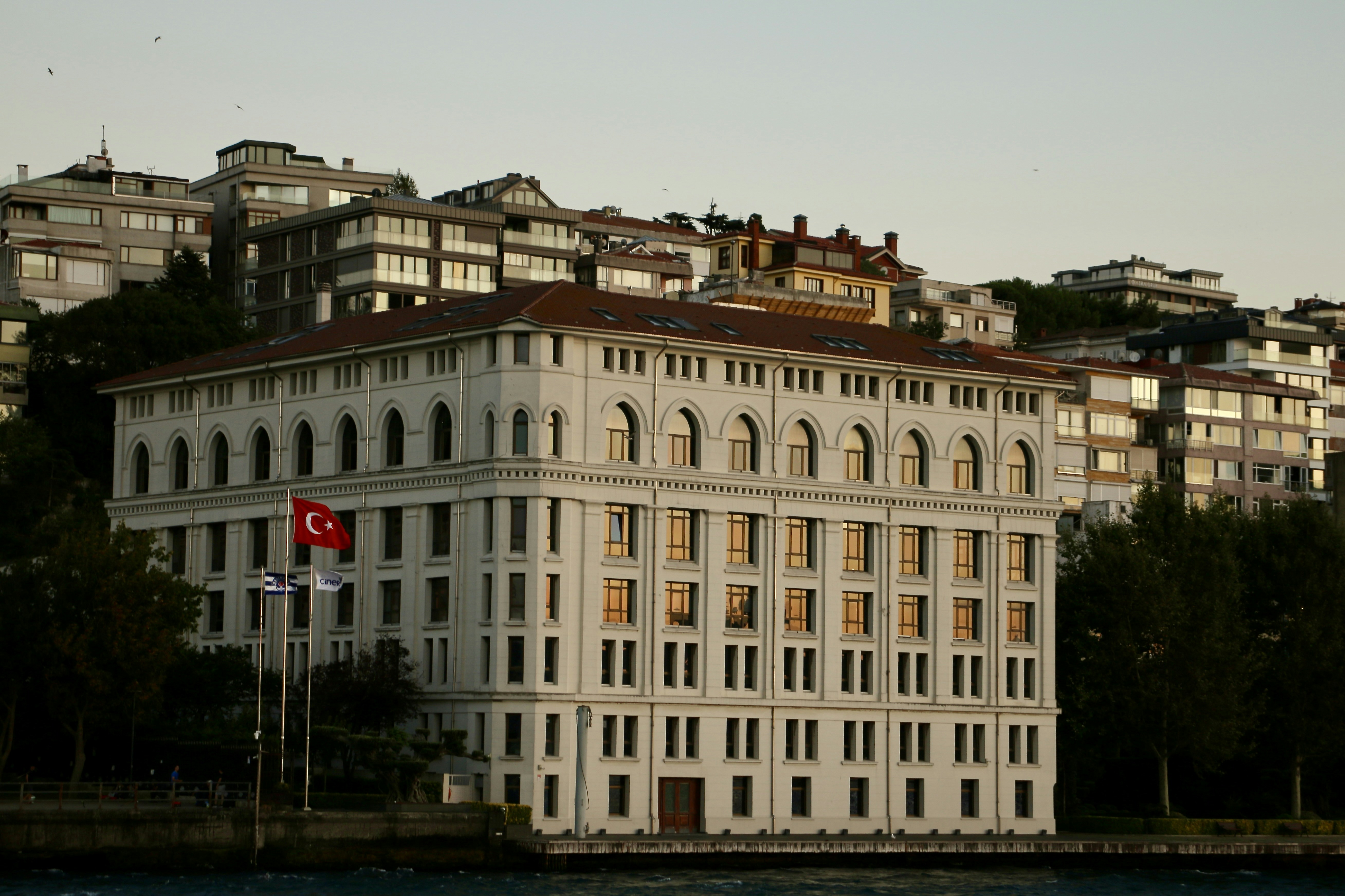 Historic building with a Turkish flag and two other flags, nestled along the Bosphorus, surrounded by modern apartments. Evening light reflects off the windows.