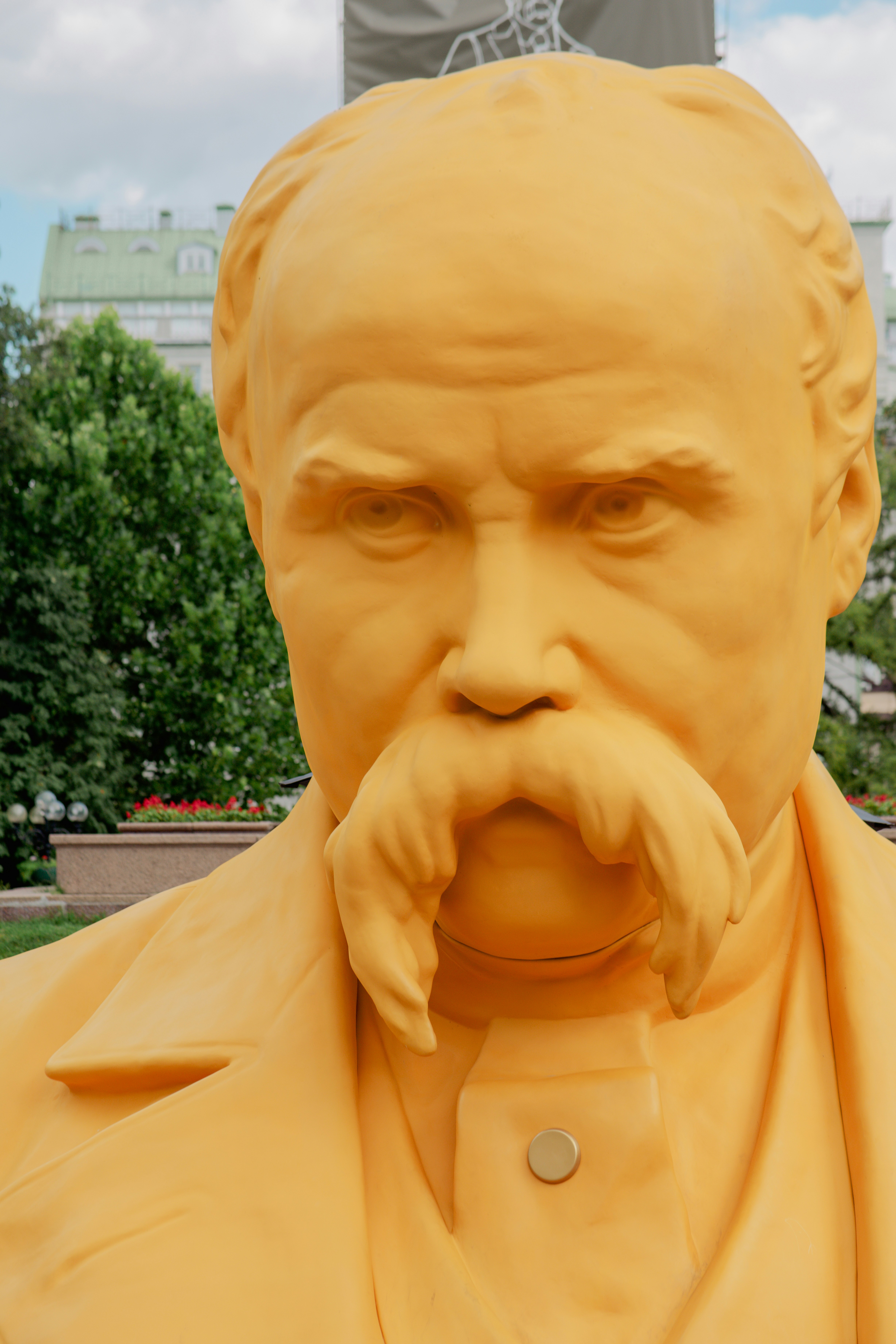 A striking yellow sculpture of a mustachioed figure, set against a backdrop of greenery and urban architecture.