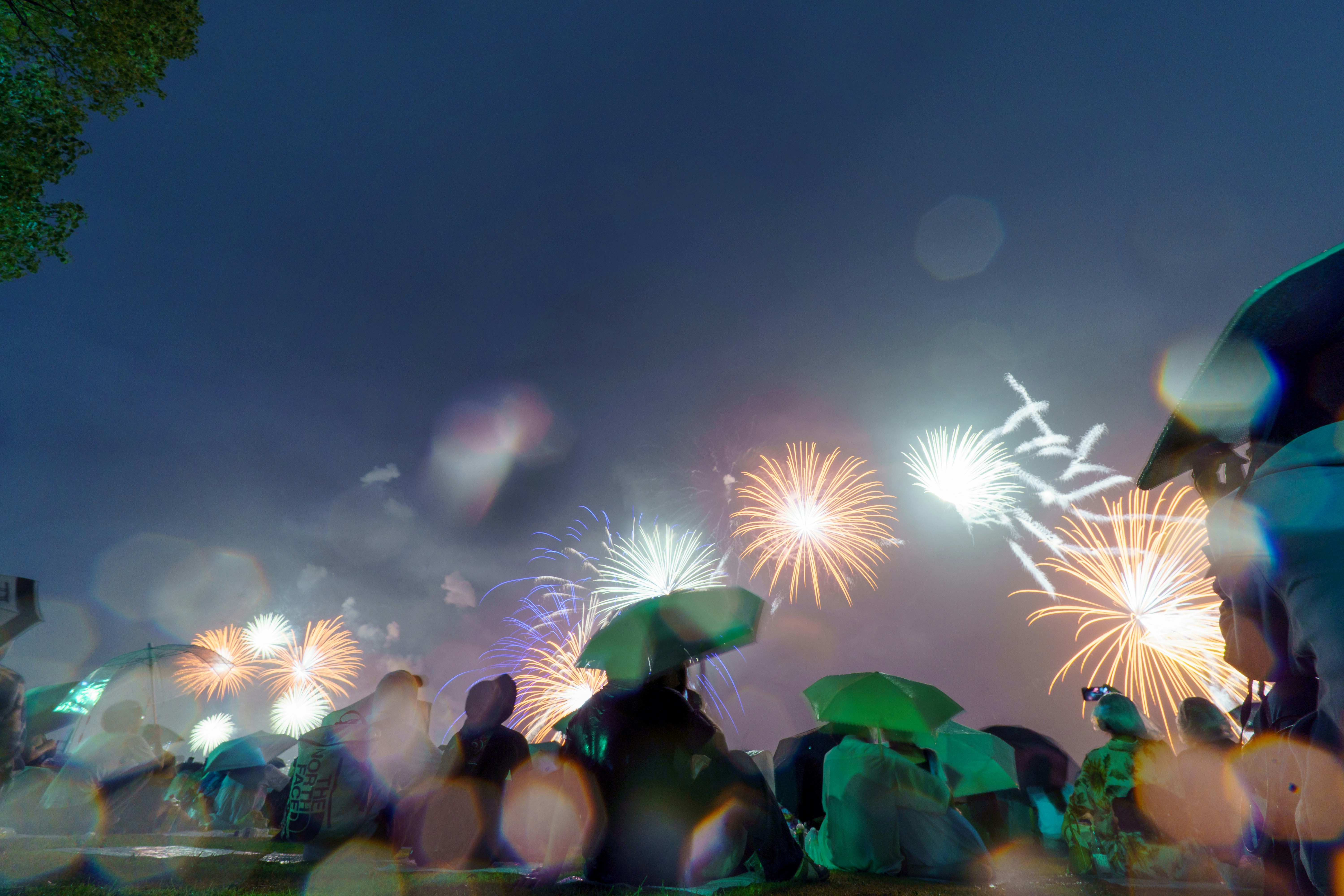 Colorful fireworks illuminate the night sky as spectators shield themselves with umbrellas, creating a vibrant atmosphere of celebration.
