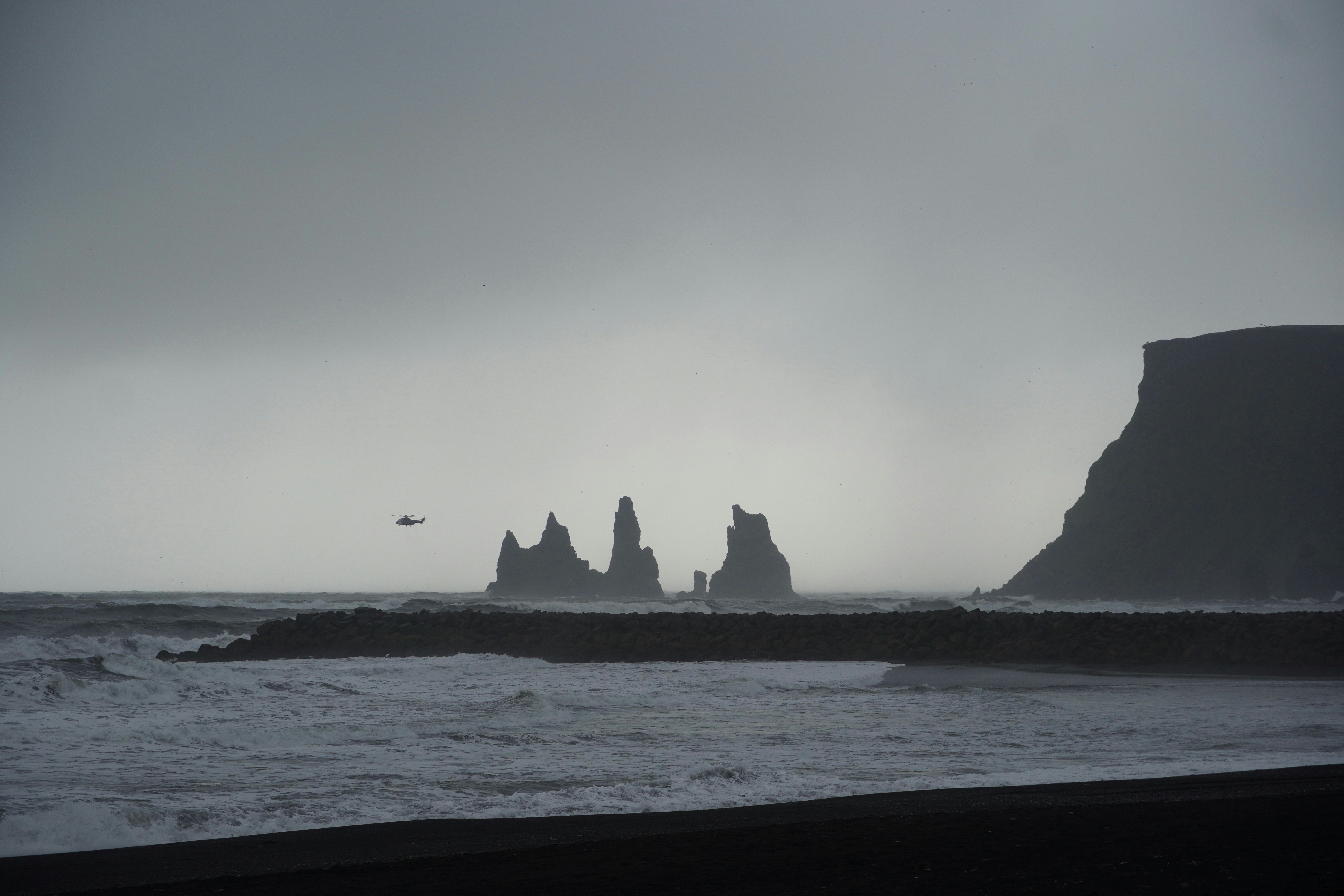 Silhouetted sea stacks rise ominously against a moody sky, with crashing waves and a distant figure adding depth to the scene.