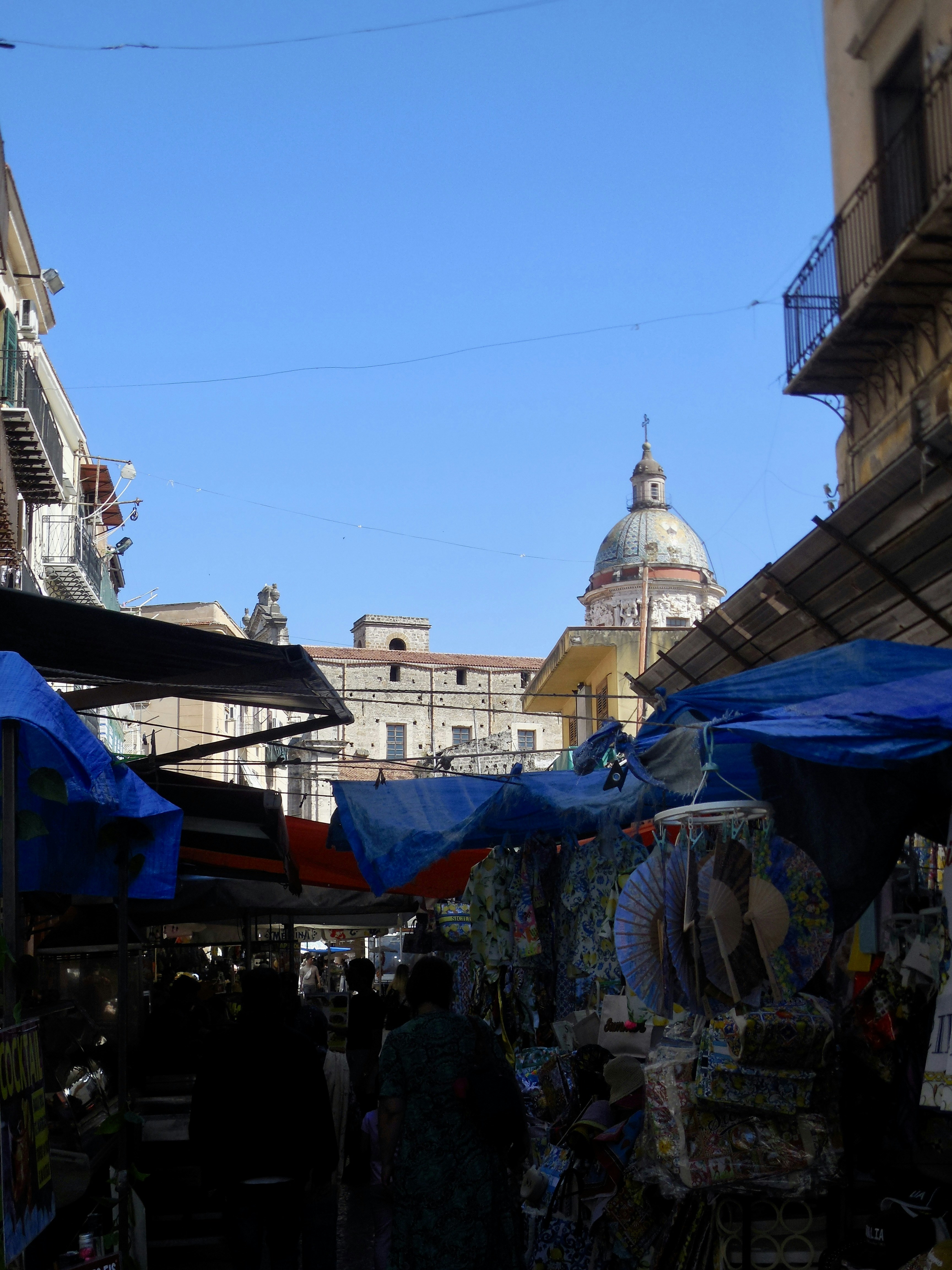 Vibrant market stalls line a narrow street, leading to a historic dome against a clear blue sky.