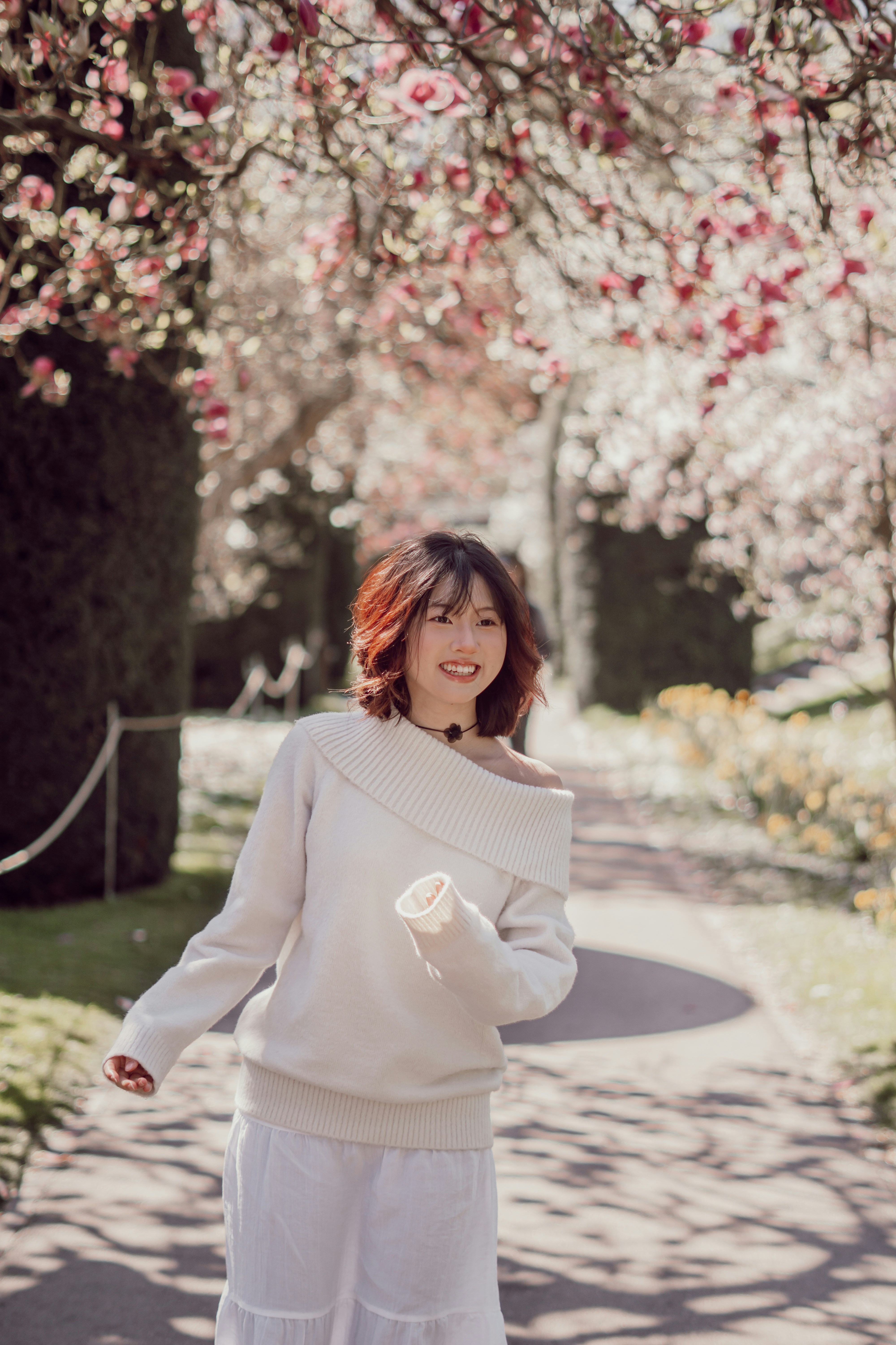 A smiling girl who is standing under a cherry tree that will blossom | A young woman in a white sweater walks outdoors.