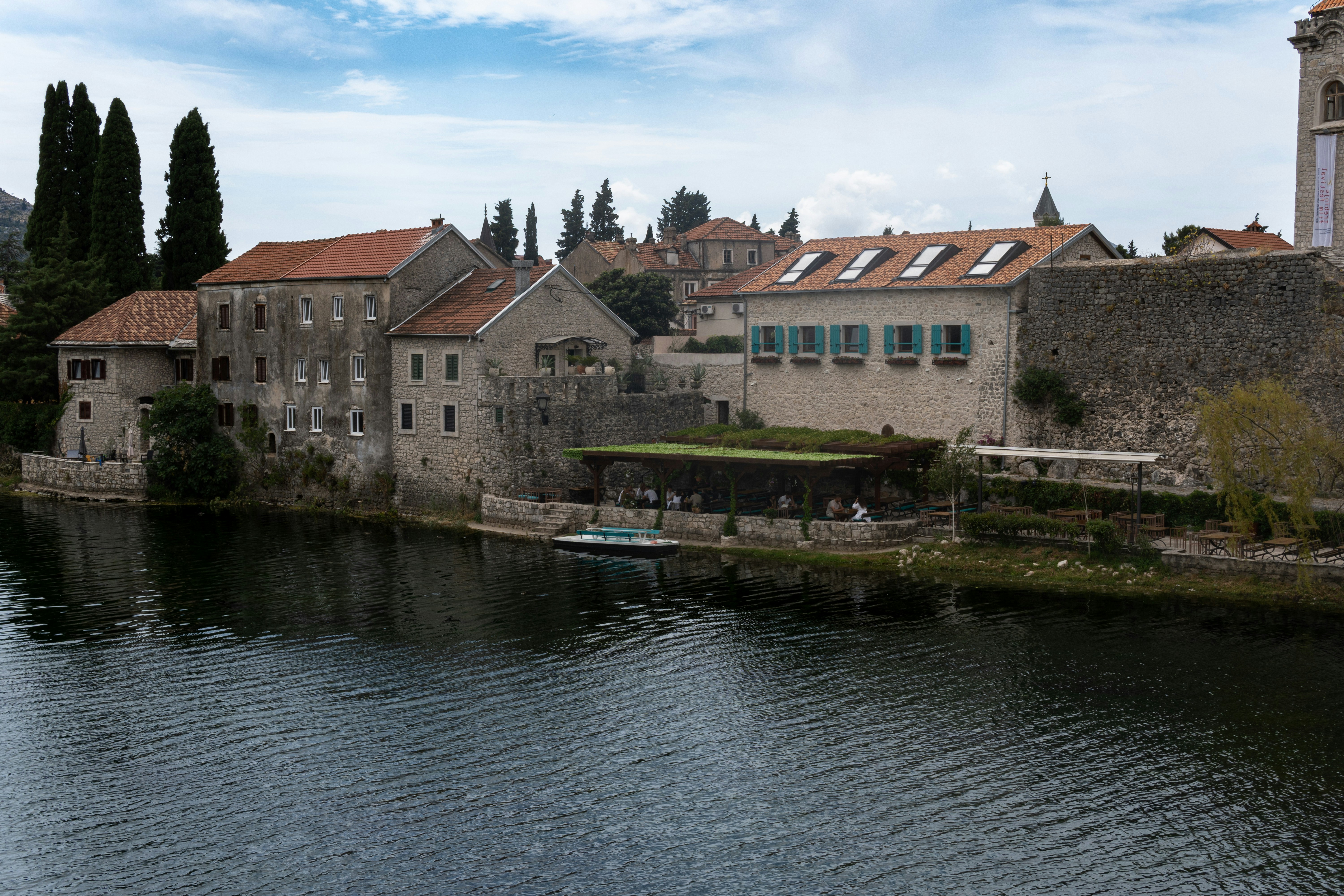Stone buildings with terracotta roofs line the riverbank, showcasing a blend of architectural styles. A wooden terrace invites visitors to enjoy the serene waterside atmosphere.