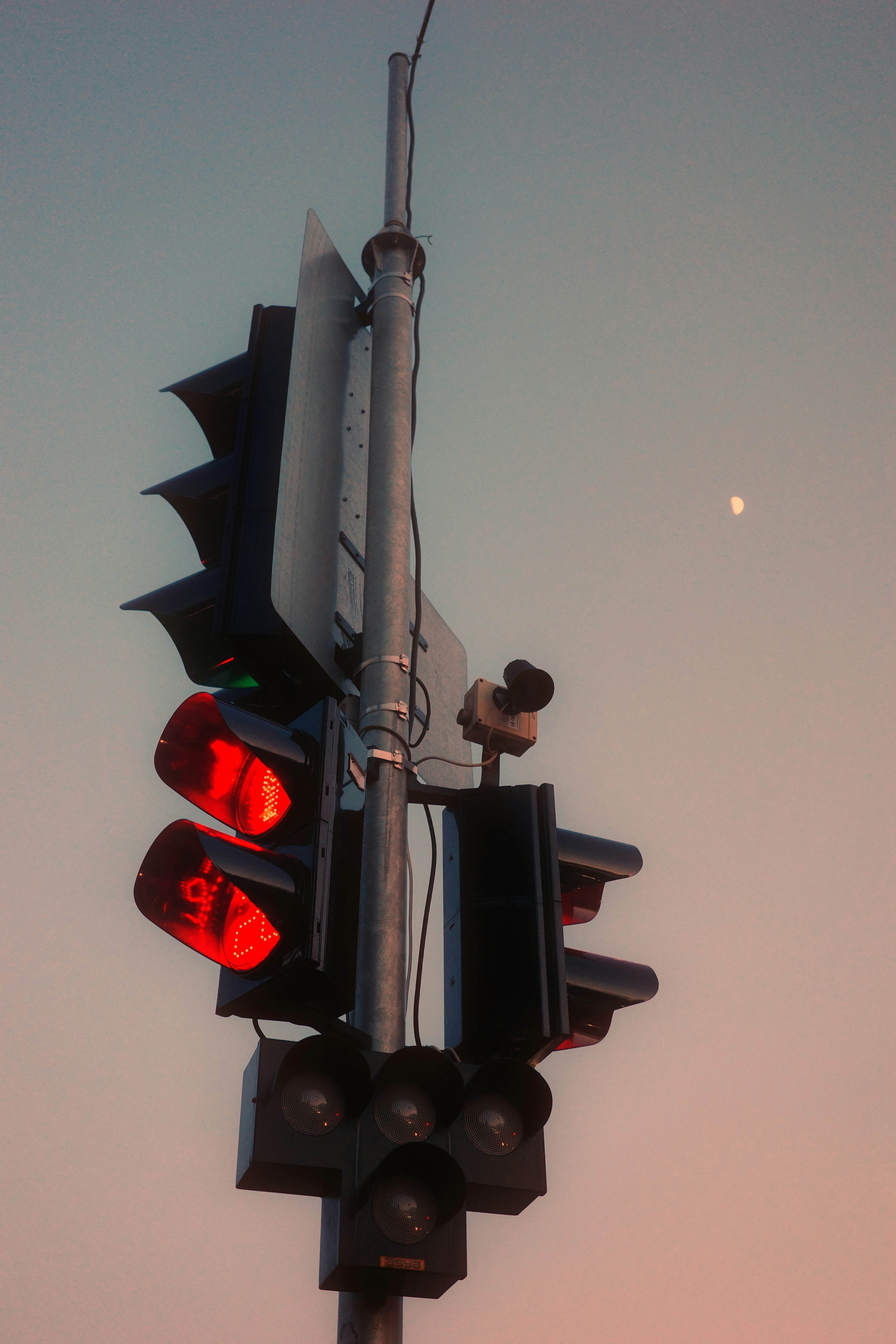 A traffic light with a lit red signal is shown against a pale, dusky sky. The street light is mounted on a tall pole, with several sets of lights visible, including a pedestrian signal below the main one. The light's metal housing is black and industrial, and wires and mounting hardware are visible. In the upper right corner of the frame, a small, crescent moon is faintly visible, offering a subtle astronomical detail that contrasts with the urban foreground. | Traffic signal showing red lights against sky