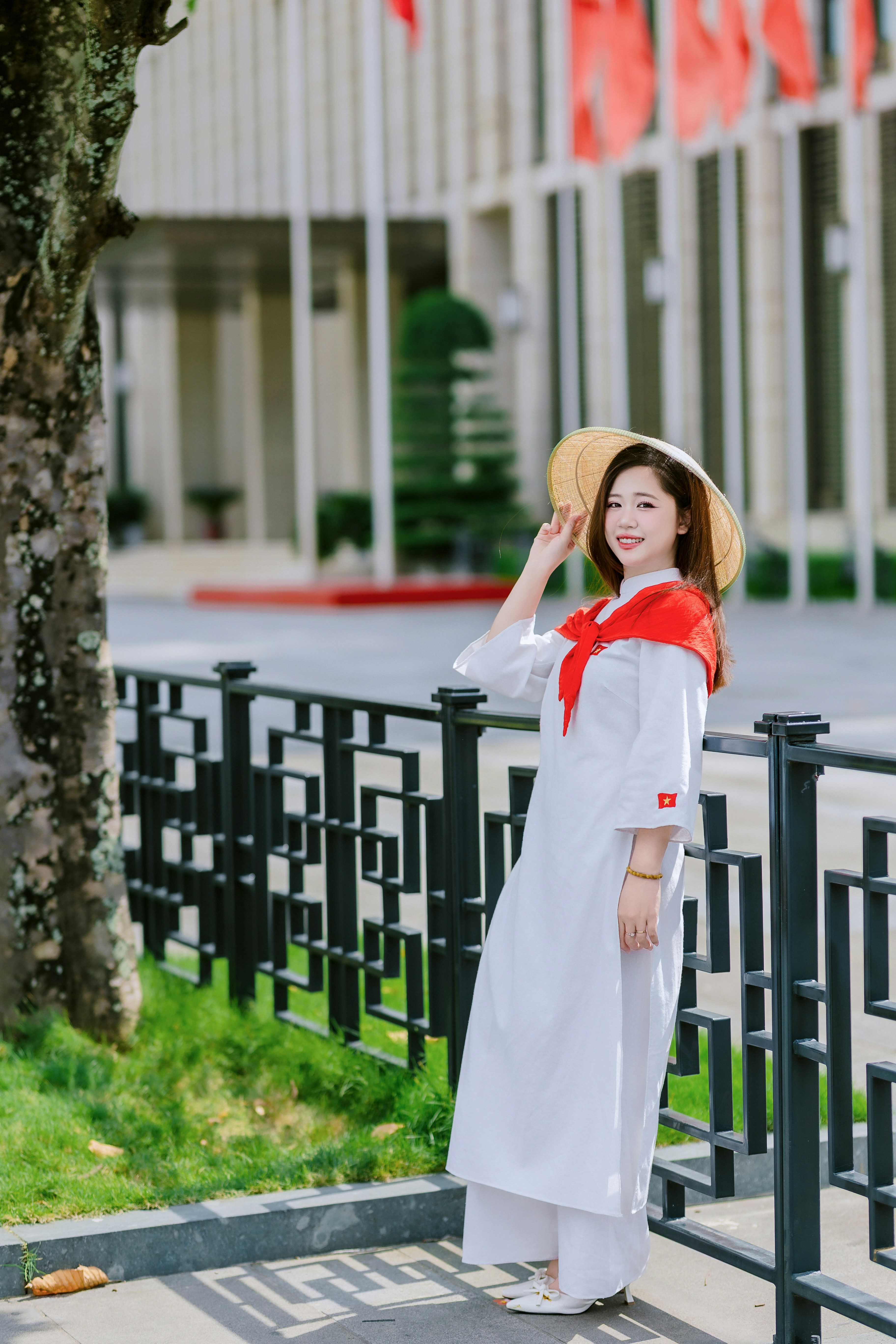 A young woman in traditional attire stands beside a decorative fence, showcasing a blend of cultural heritage and contemporary surroundings.