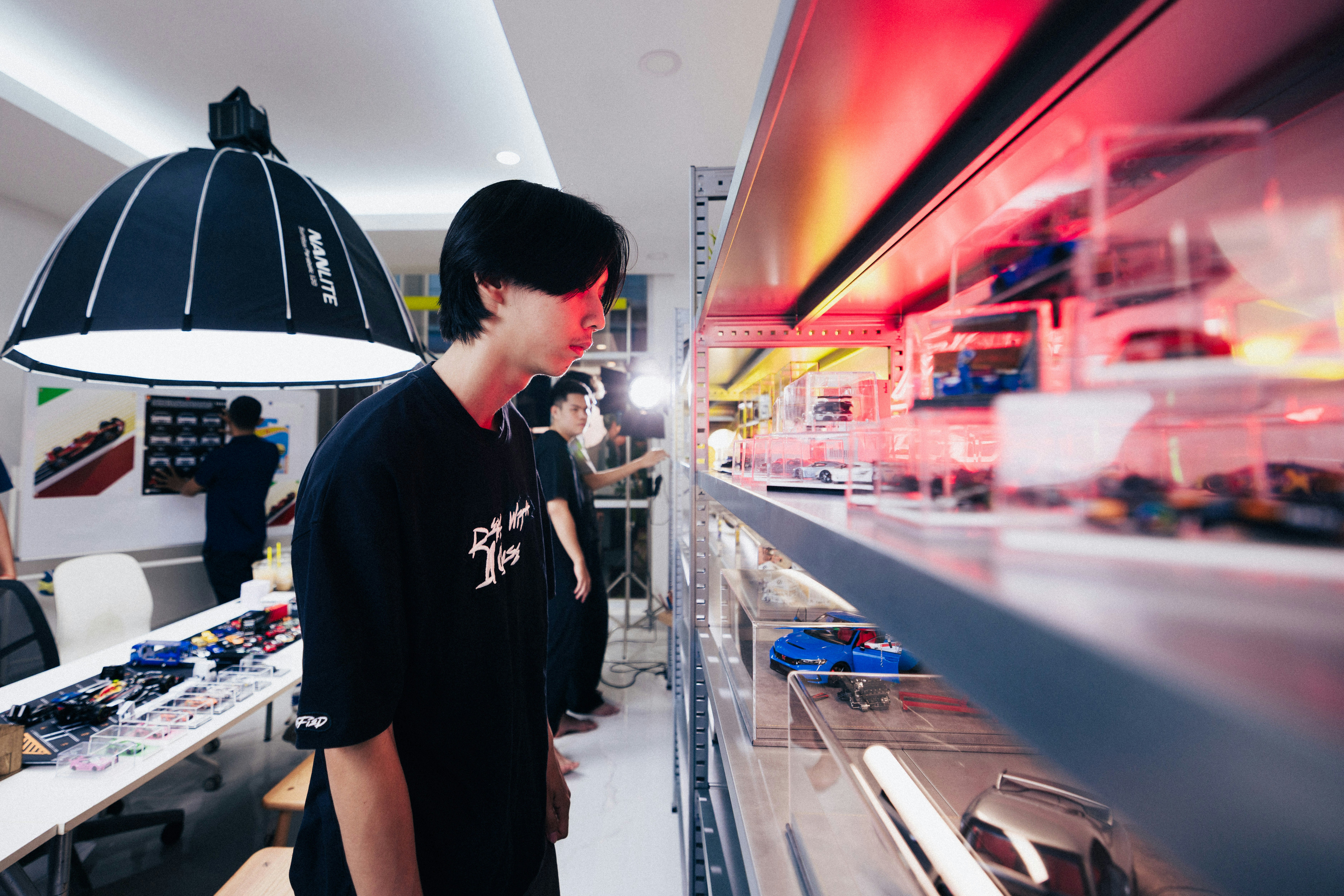 A young man examines a display of miniature cars under vibrant lighting in a creative workspace. The scene showcases the intersection of art and hobby.
