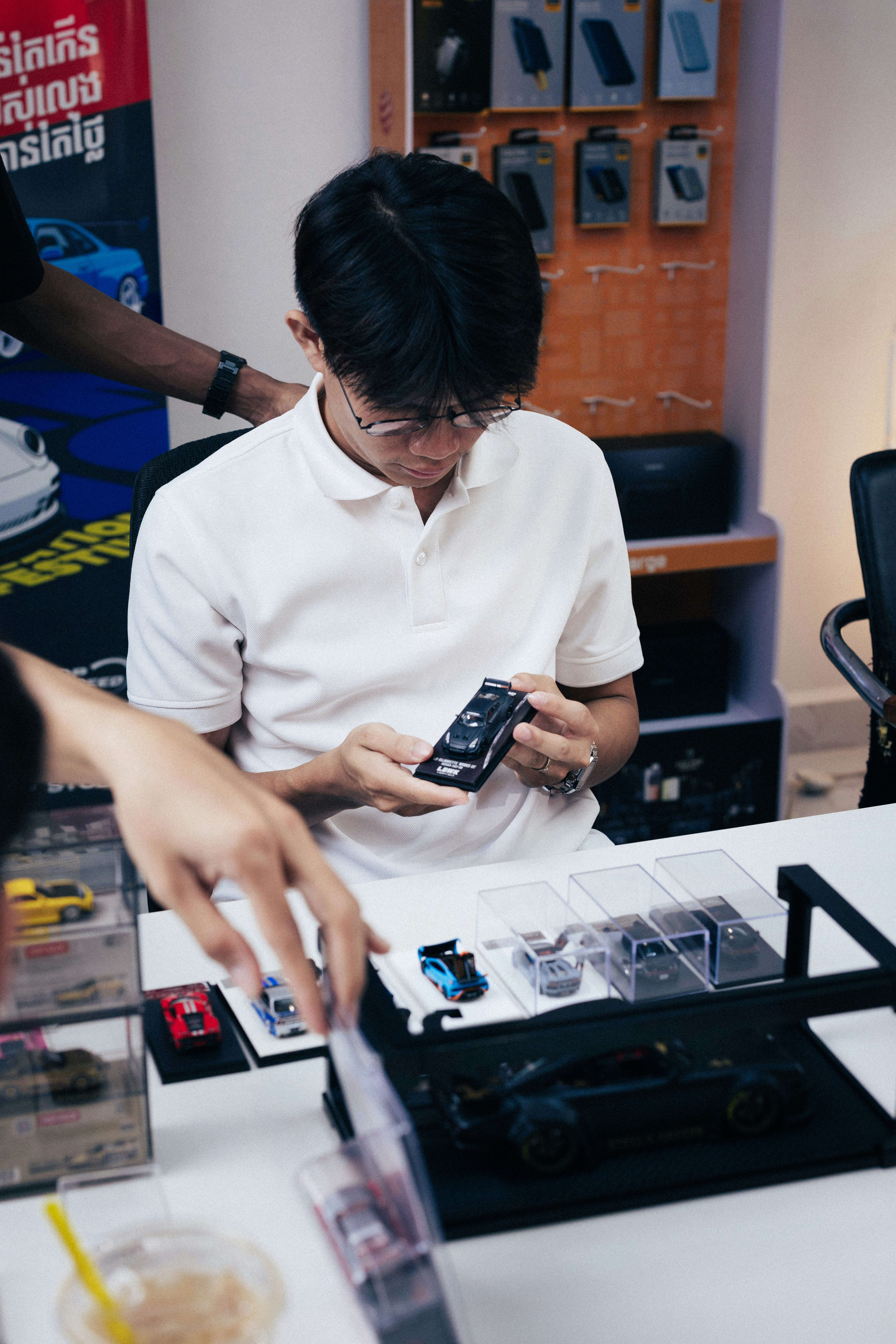 A young man examines a detailed model car while surrounded by various miniature vehicles displayed in clear cases. The scene captures the passion of a collector's hobby.