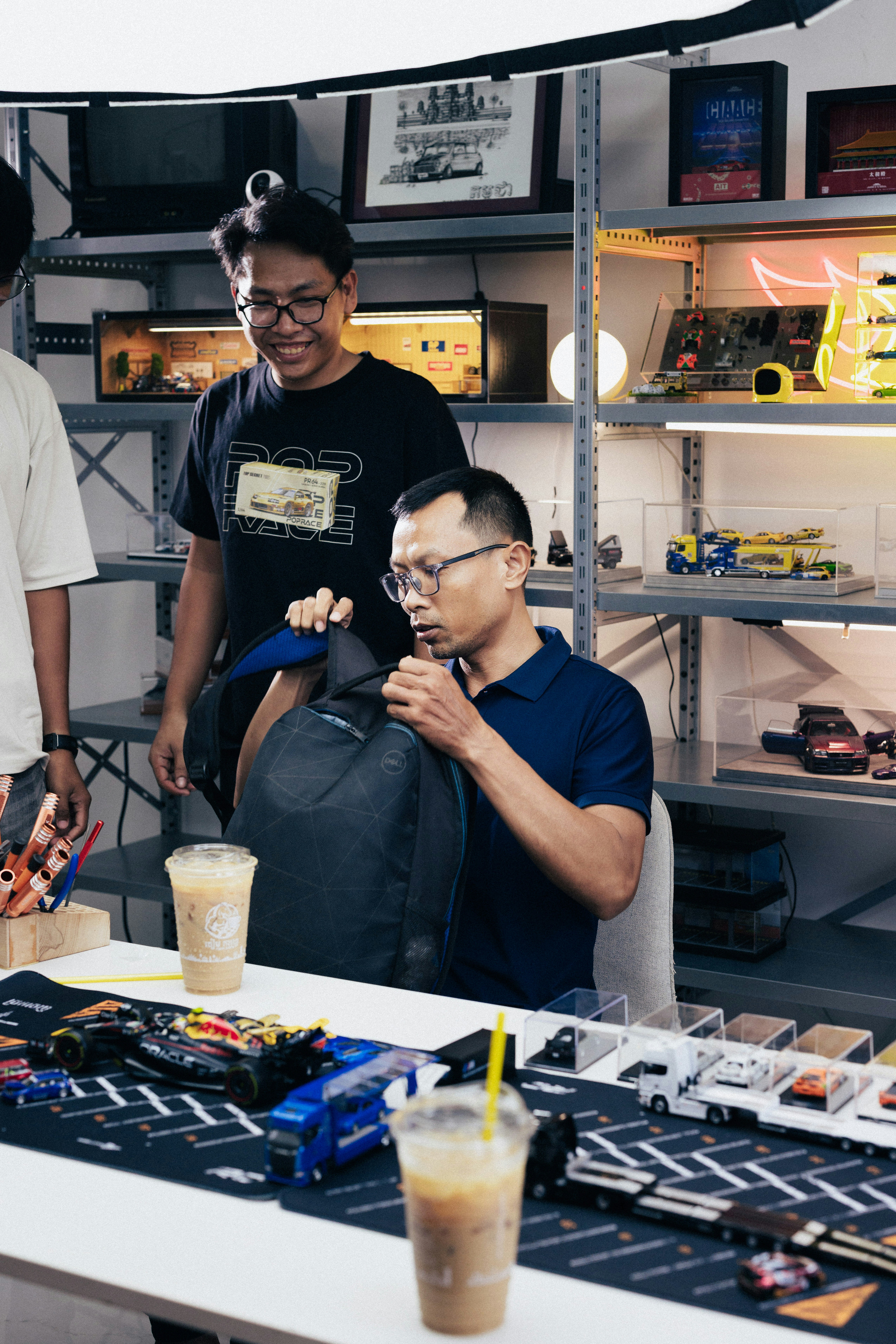 Three individuals engage in a creative session, examining miniature vehicles on a table while discussing ideas. A backdrop of collectibles adds to the atmosphere.