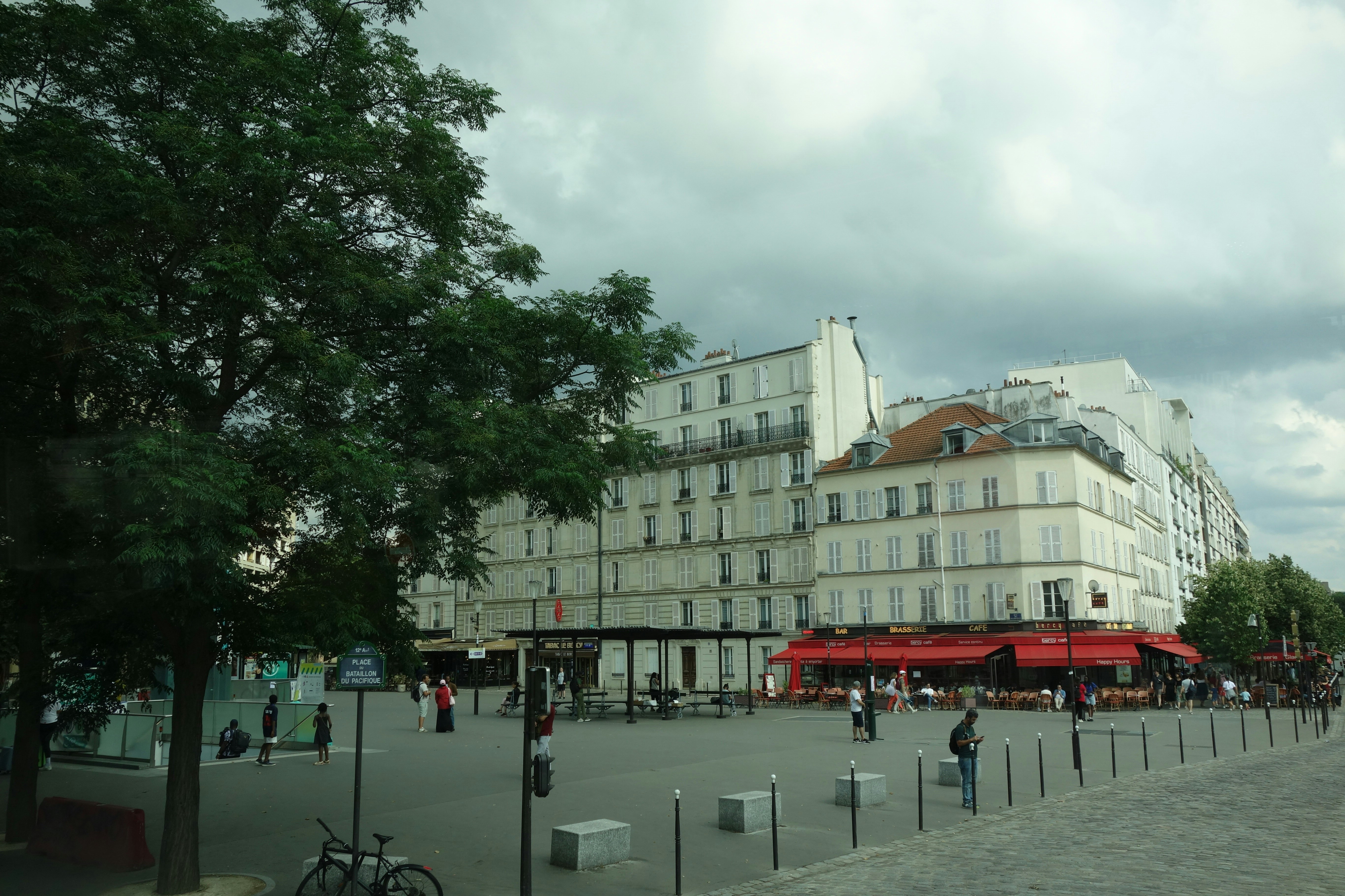 A bustling Parisian square with people mingling, surrounded by classic architecture and shaded by lush trees. The vibrant café scene adds a lively touch.
