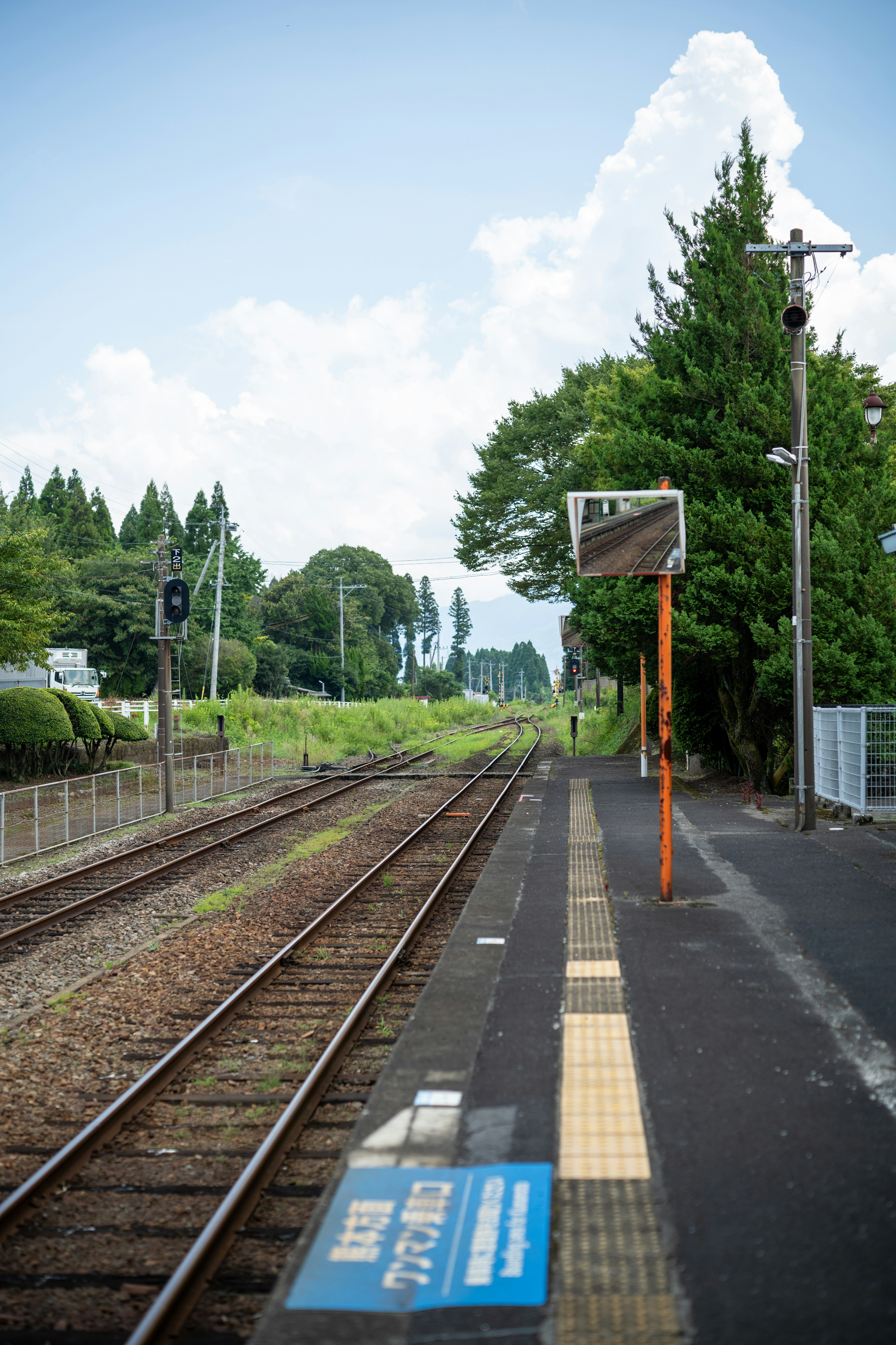 Abandoned train station platform with overgrown tracks, surrounded by lush greenery and a clear blue sky. A mirror reflects the serene landscape.