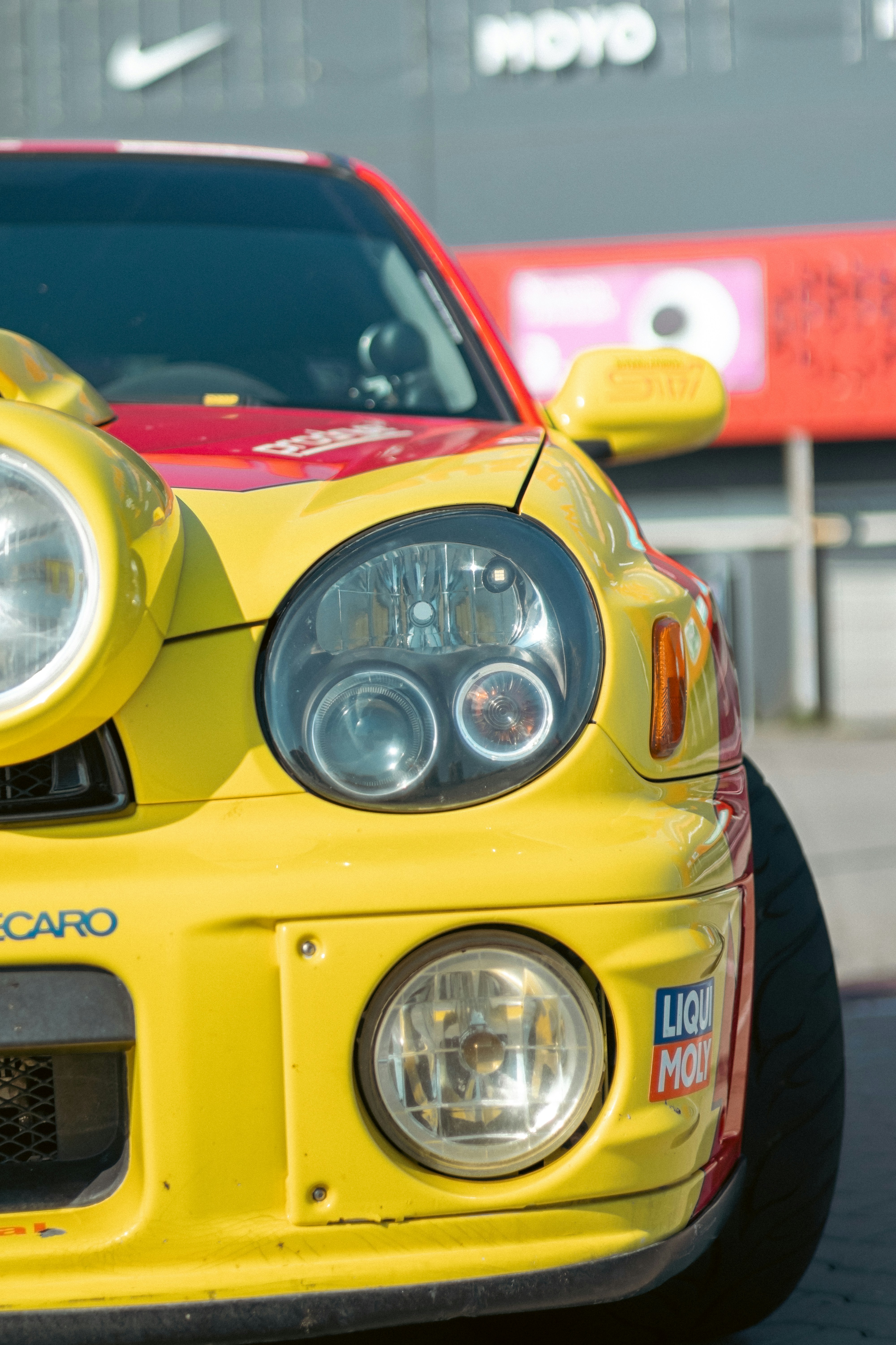 Close-up view of a brightly colored race car's front end, showcasing its headlights and aggressive styling. The background features branding elements that enhance the racing theme.
