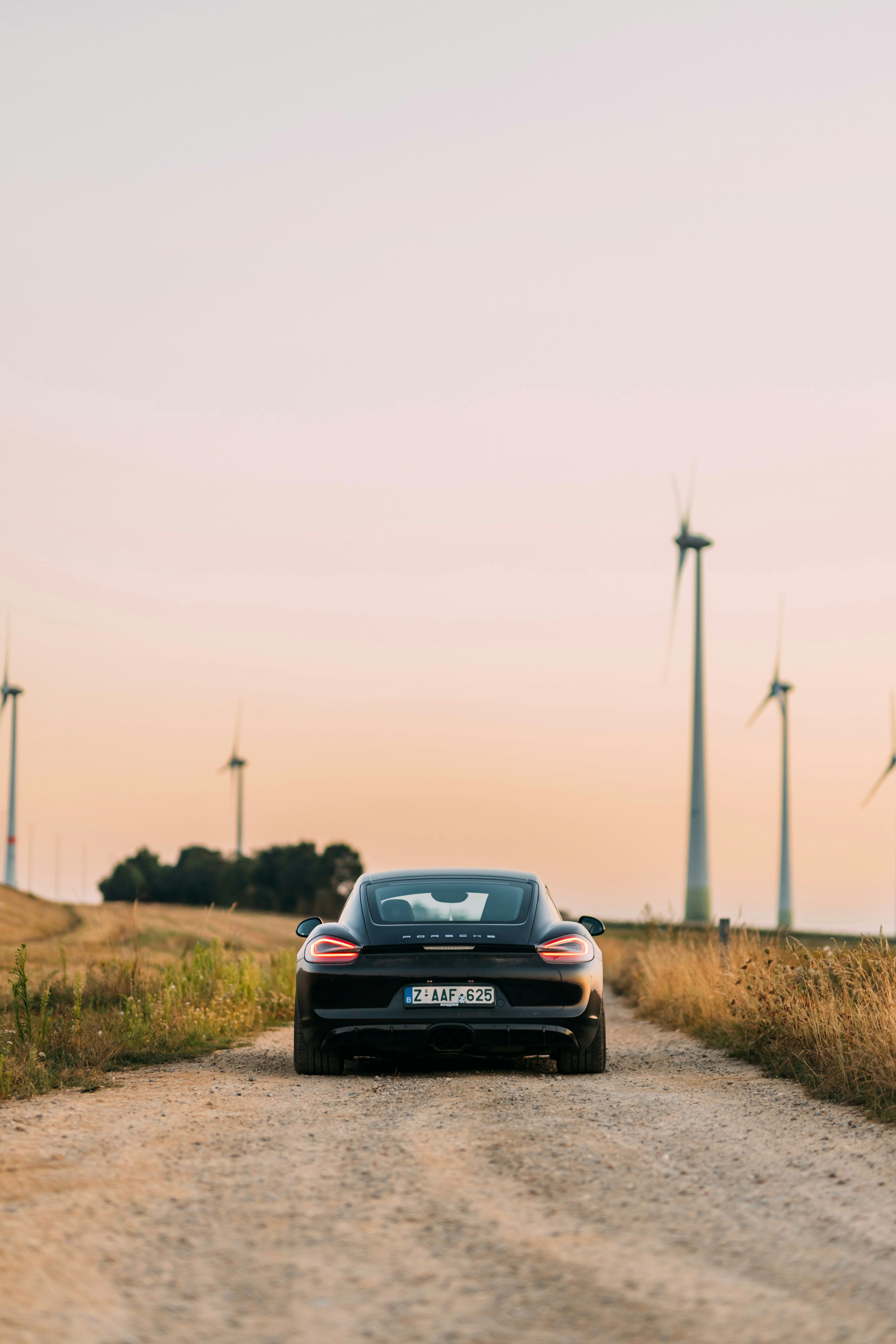 A sleek black Porsche parked on a gravel road, framed by tall wind turbines against a pastel sunset sky.