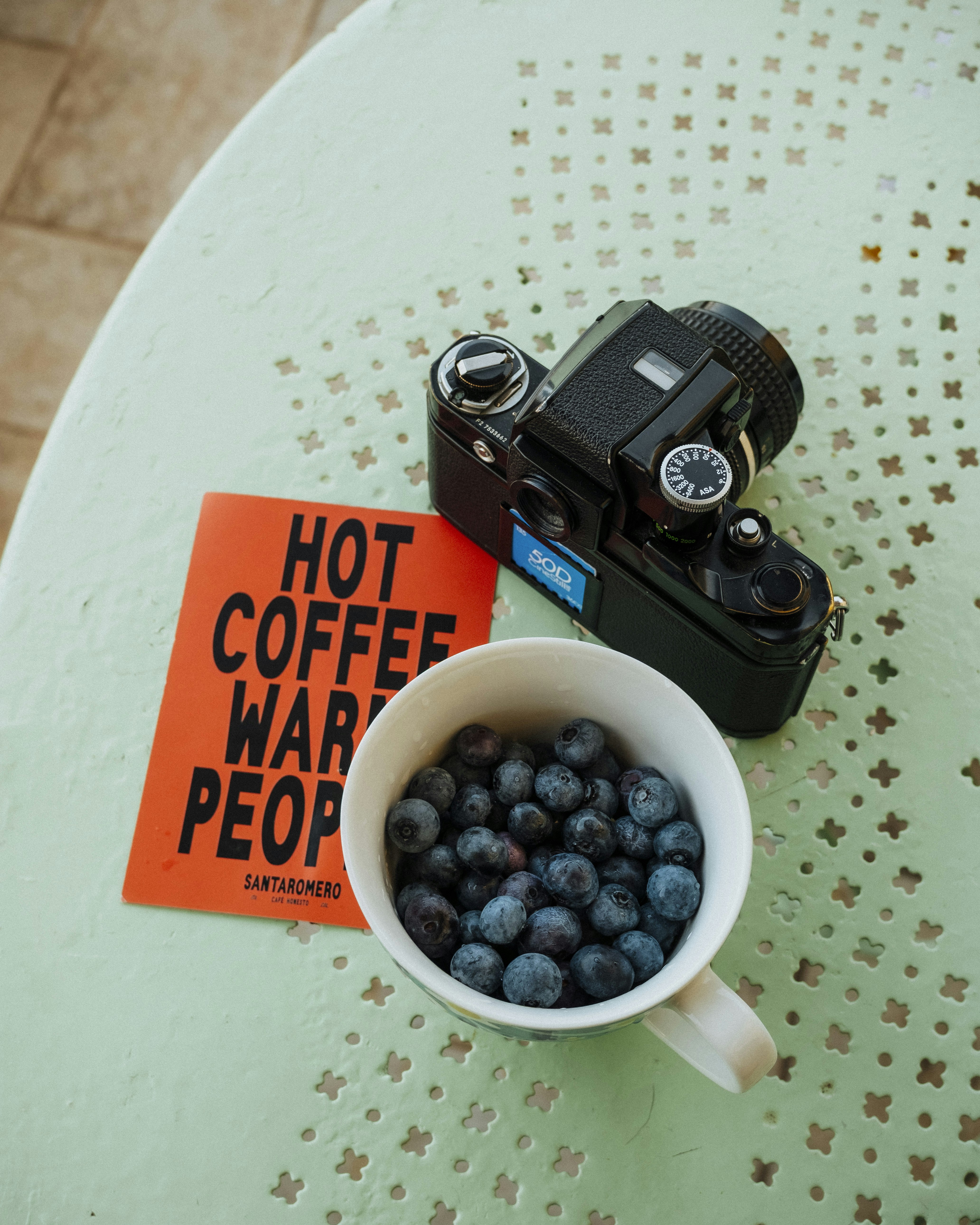 A vintage camera rests beside a cup filled with fresh blueberries on a textured table, accompanied by a bold orange book cover. 