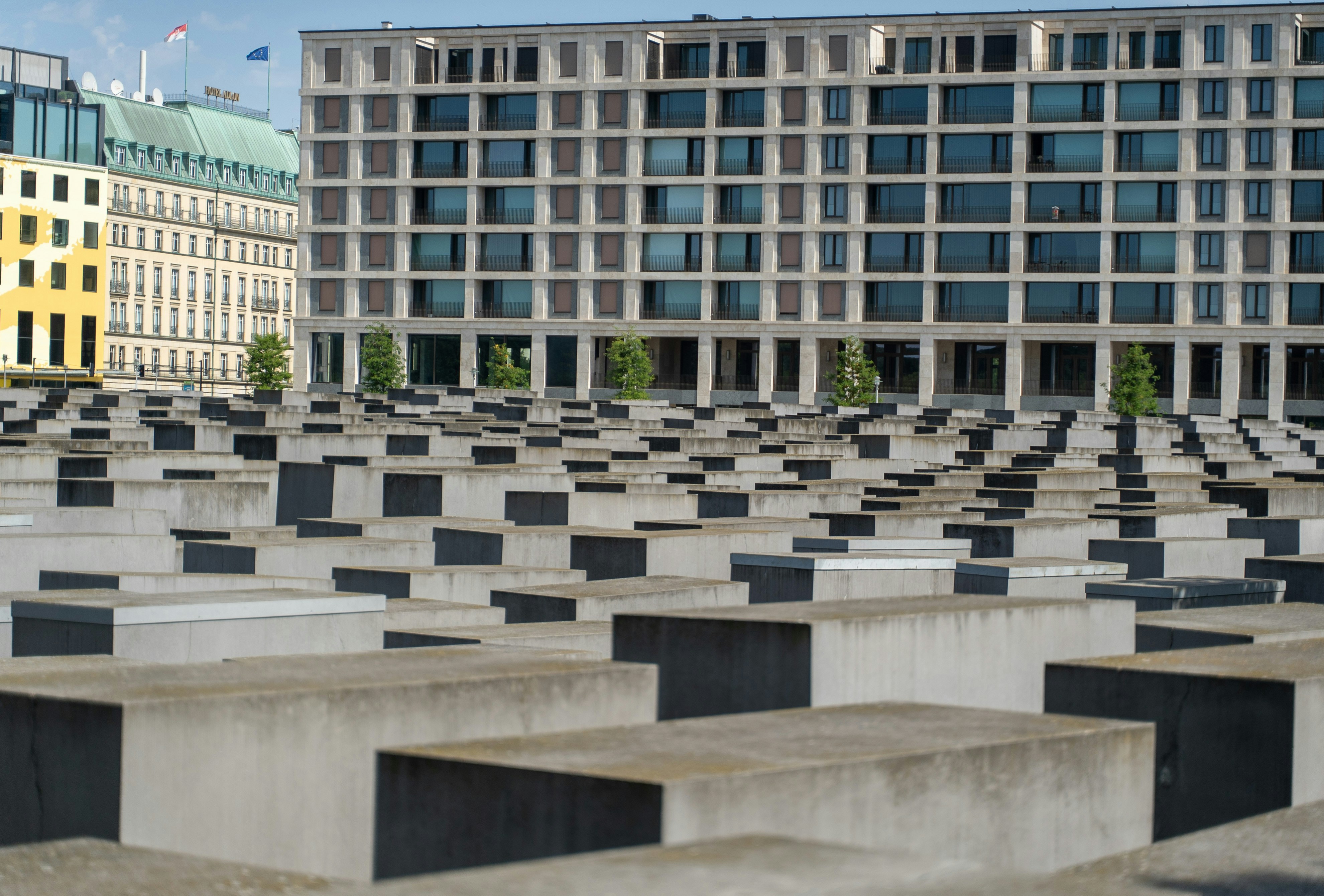 Abstract arrangement of concrete blocks in a memorial site, juxtaposed against contemporary architecture and greenery.