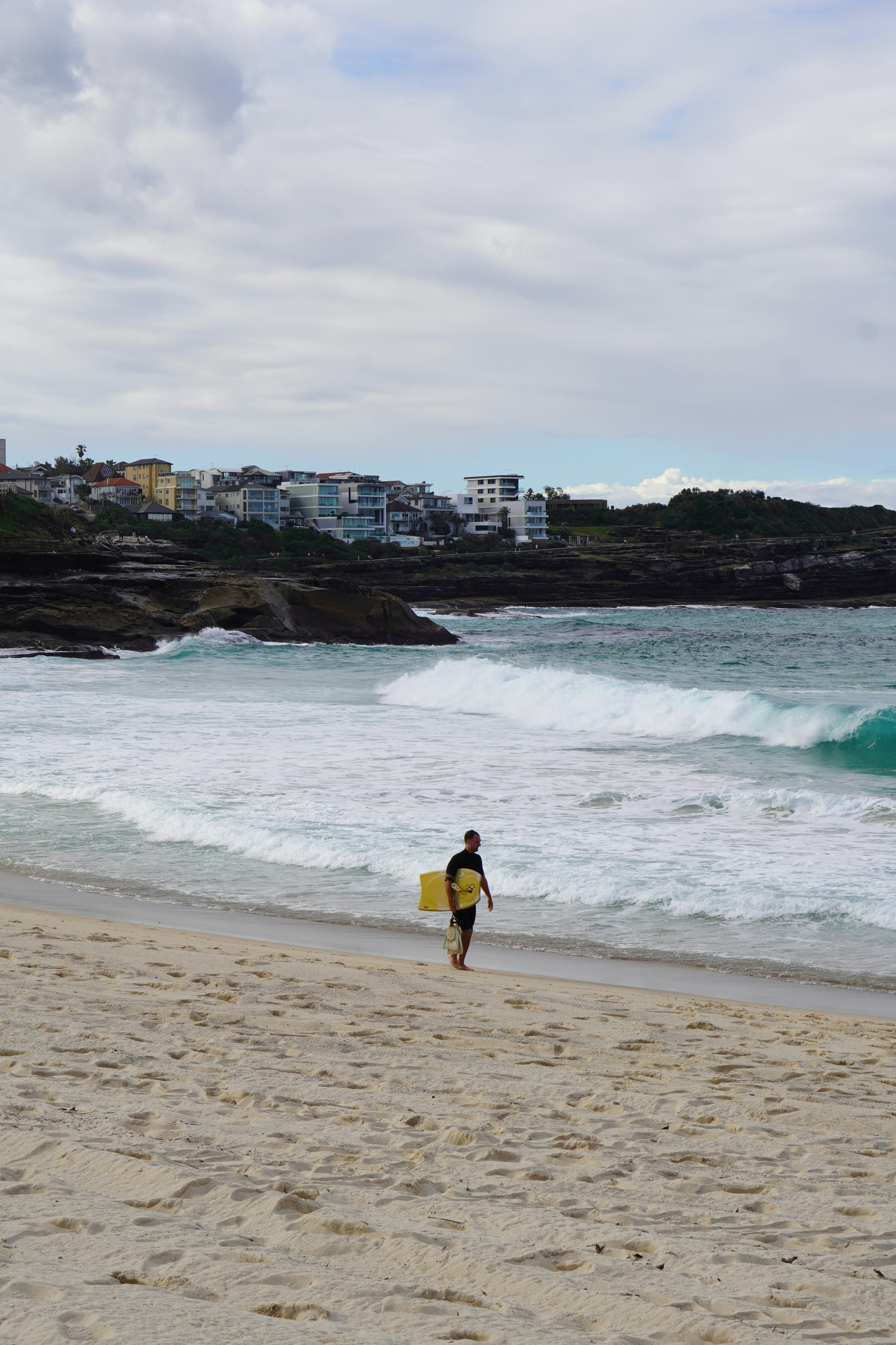 Surfer walking along the sandy beach with a yellow board, waves crashing in the background and coastal buildings visible.