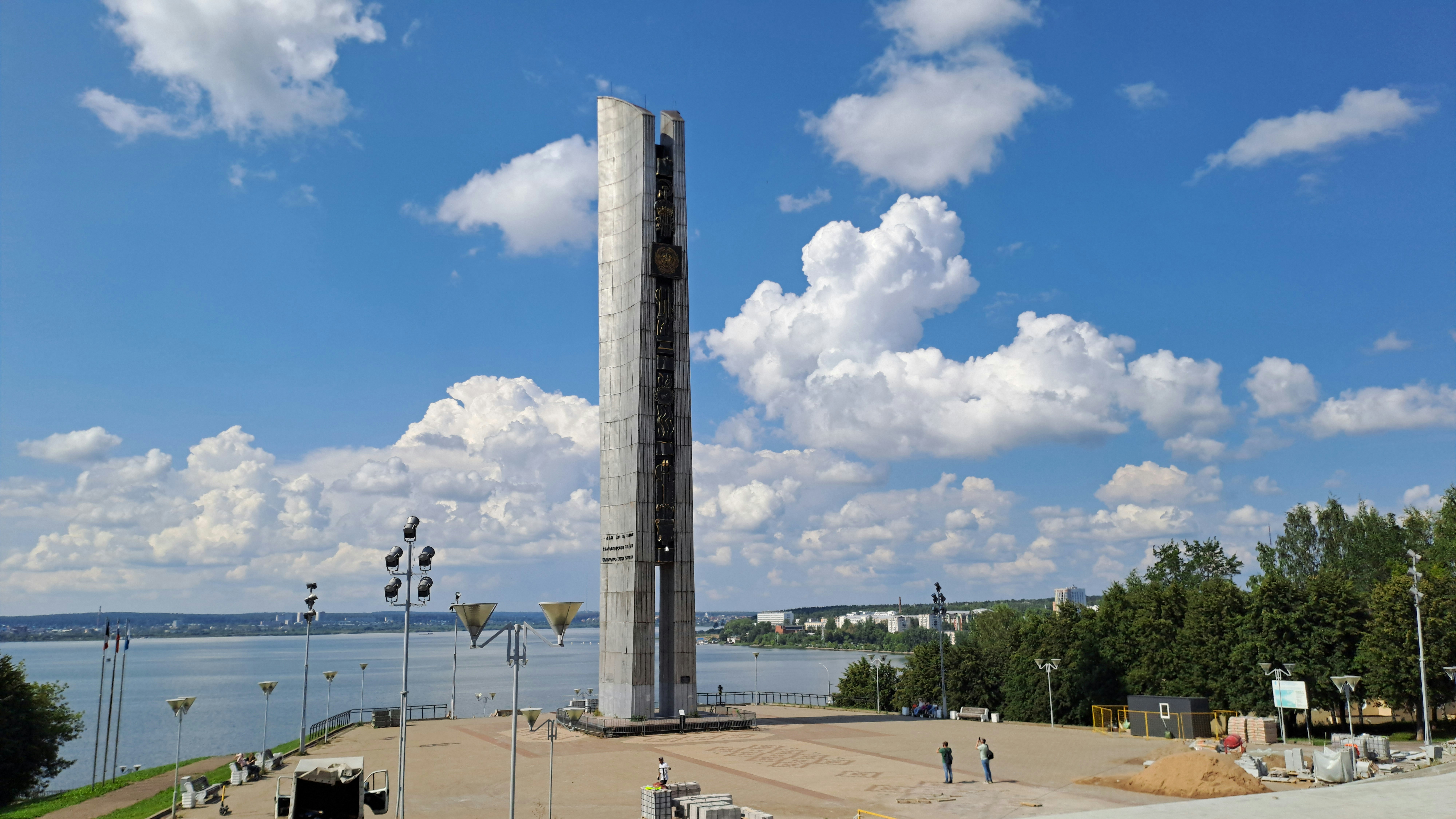 The Izhevsk Esplanade is an urban development ensemble in the Oktyabrsky District of the city in the form of a pedestrian zone consisting of several boulevards and squares connecting the Zodchy Dudin Embankment in the west and the Central Square in the east. The monumental dominant in the west is the Friendship of Peoples monument, in the east the ensemble is closed by the building of the Government House of the Udmurt Republic. The Esplanade is a recognizable symbol of Izhevsk and determines the appearance of its central part. | Tall modern sculpture beside a body of water