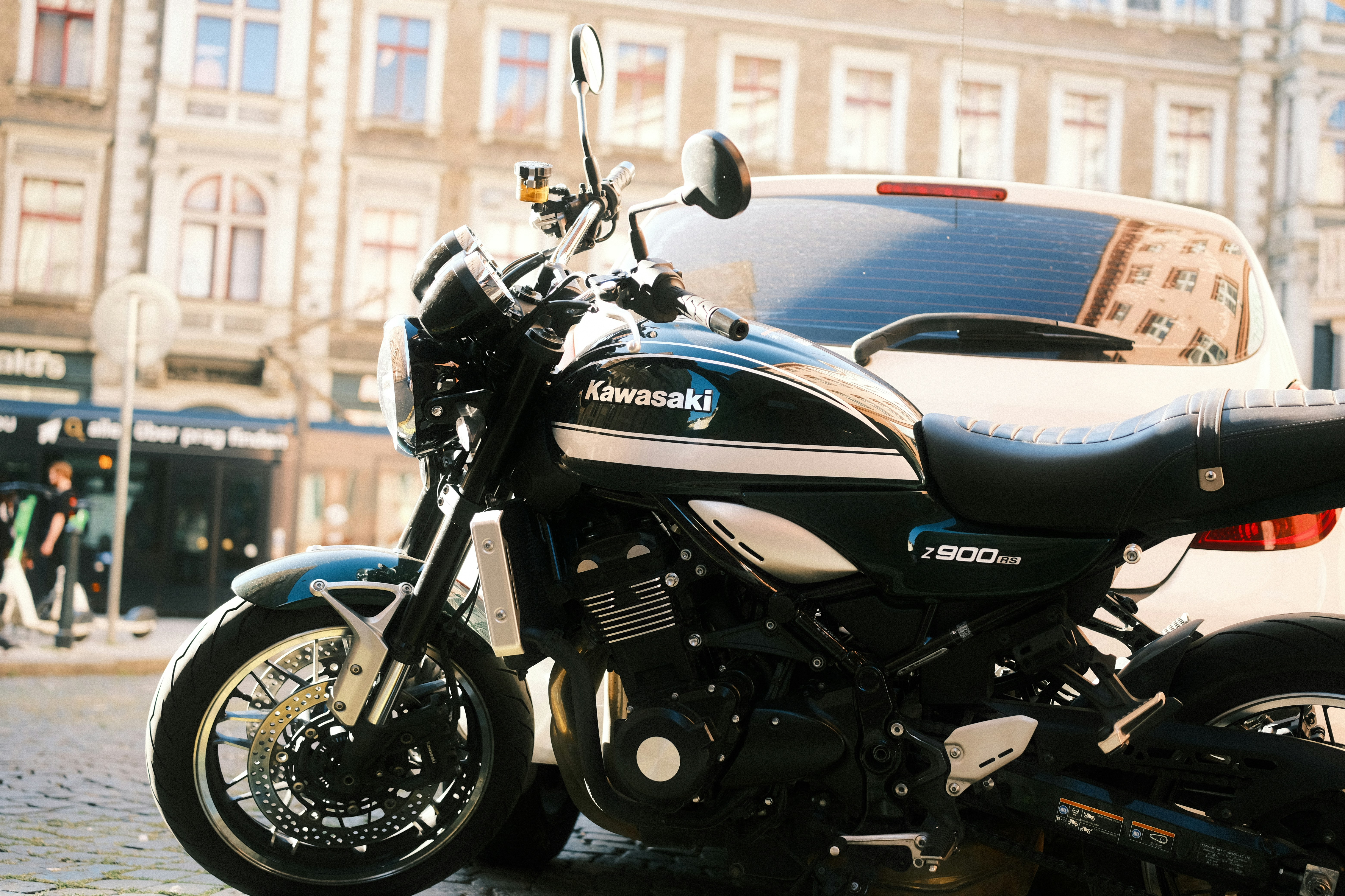Kawasaki Z900RS. A green Kawasaki motorcycle is parked beside a white vehicle on a cobblestone street. Buildings and pedestrians are visible in the background during daylight.