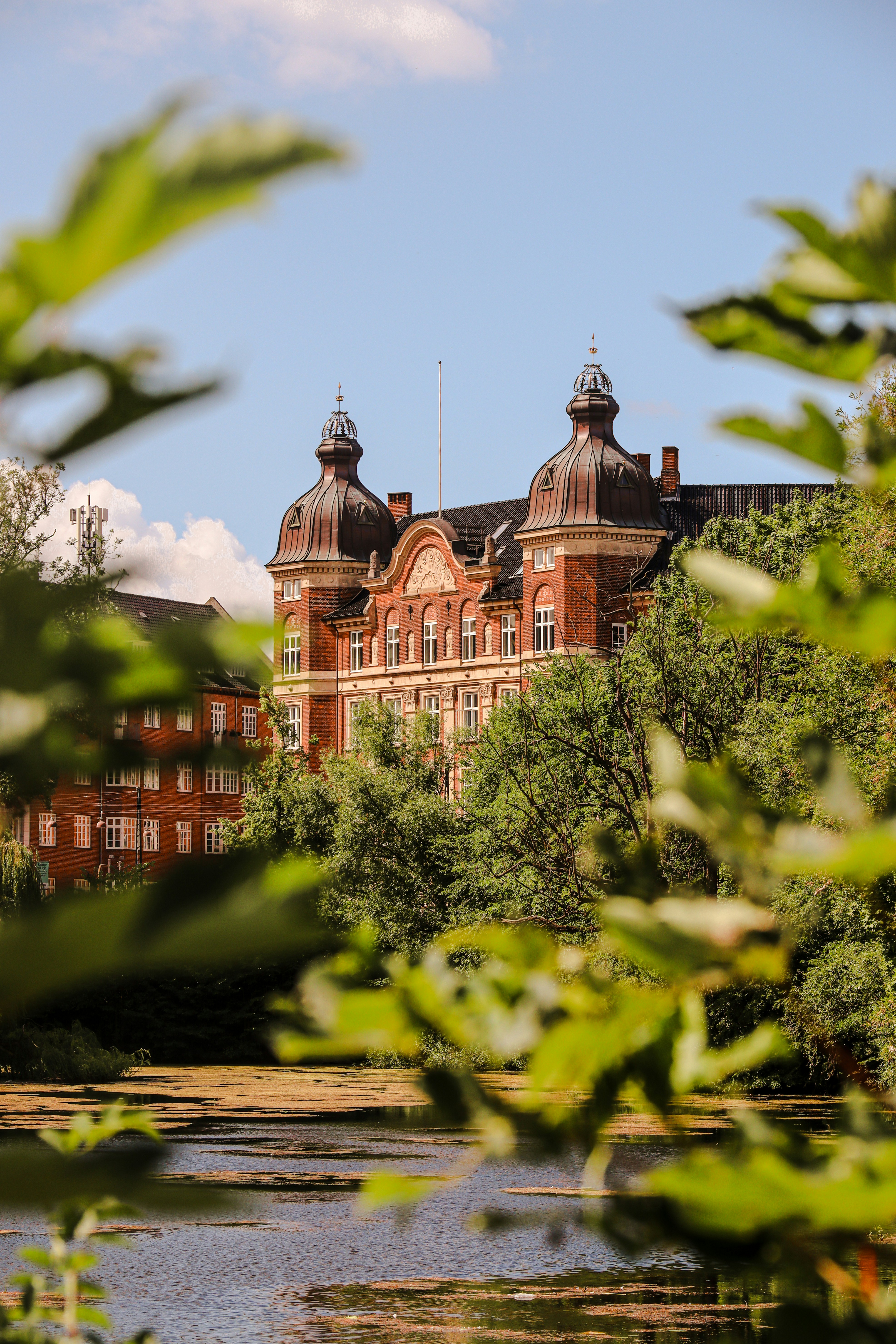 Historic building framed by greenery near a serene lake in Copenhagen | Historic building seen through green foliage and water.
