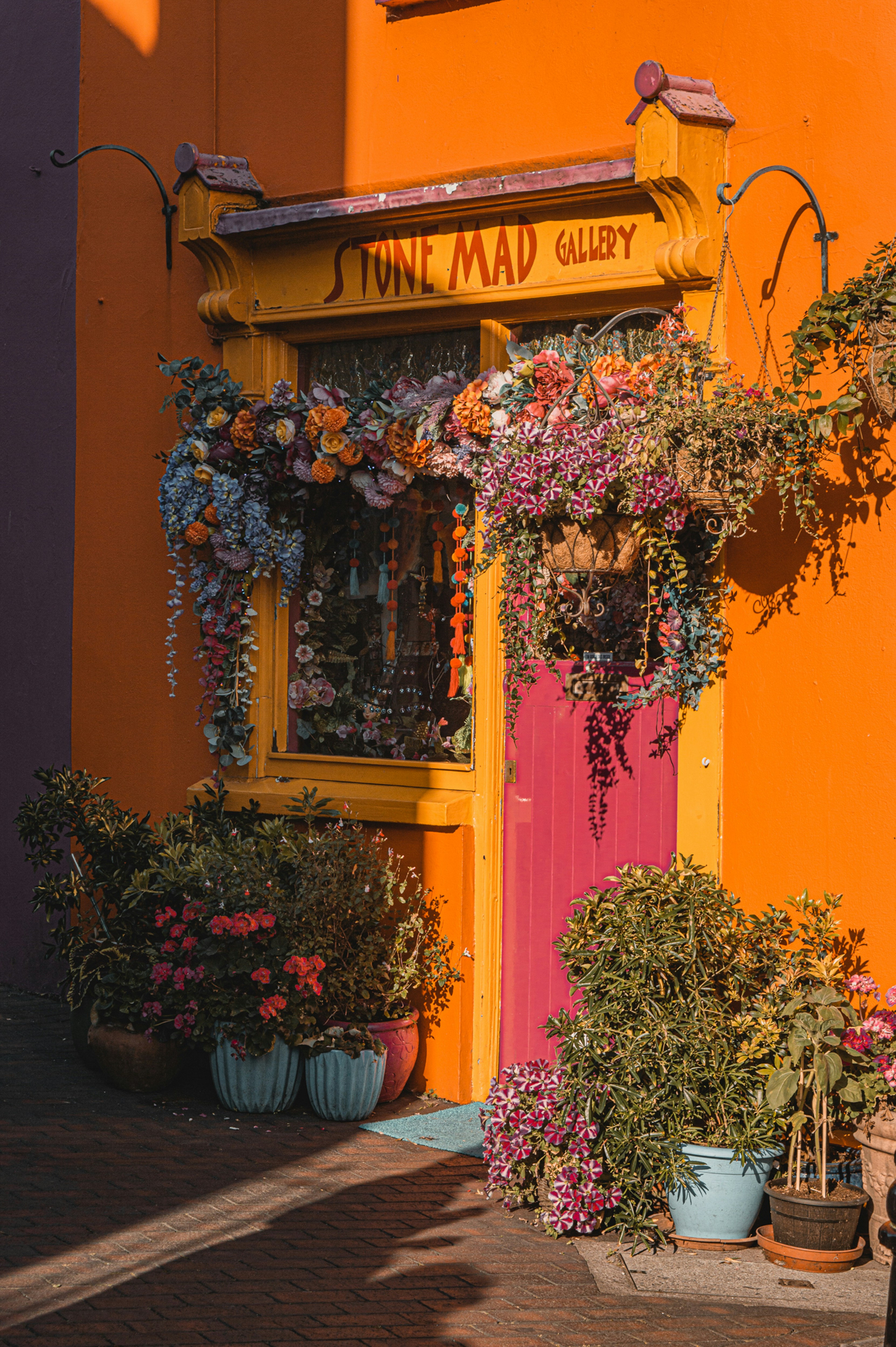 Kinsale is a vibrant coastal town renowned for its colorful streets, lively culinary scene, and breathtaking harbor views, making it ideal to visit in summer. Visitors can enjoy walking tours, explore historic sites like Charles Fort and James Fort, or take to the sea with sailing, kayaking, and harbor cruises. | Brightly painted shopfront with abundant floral displays