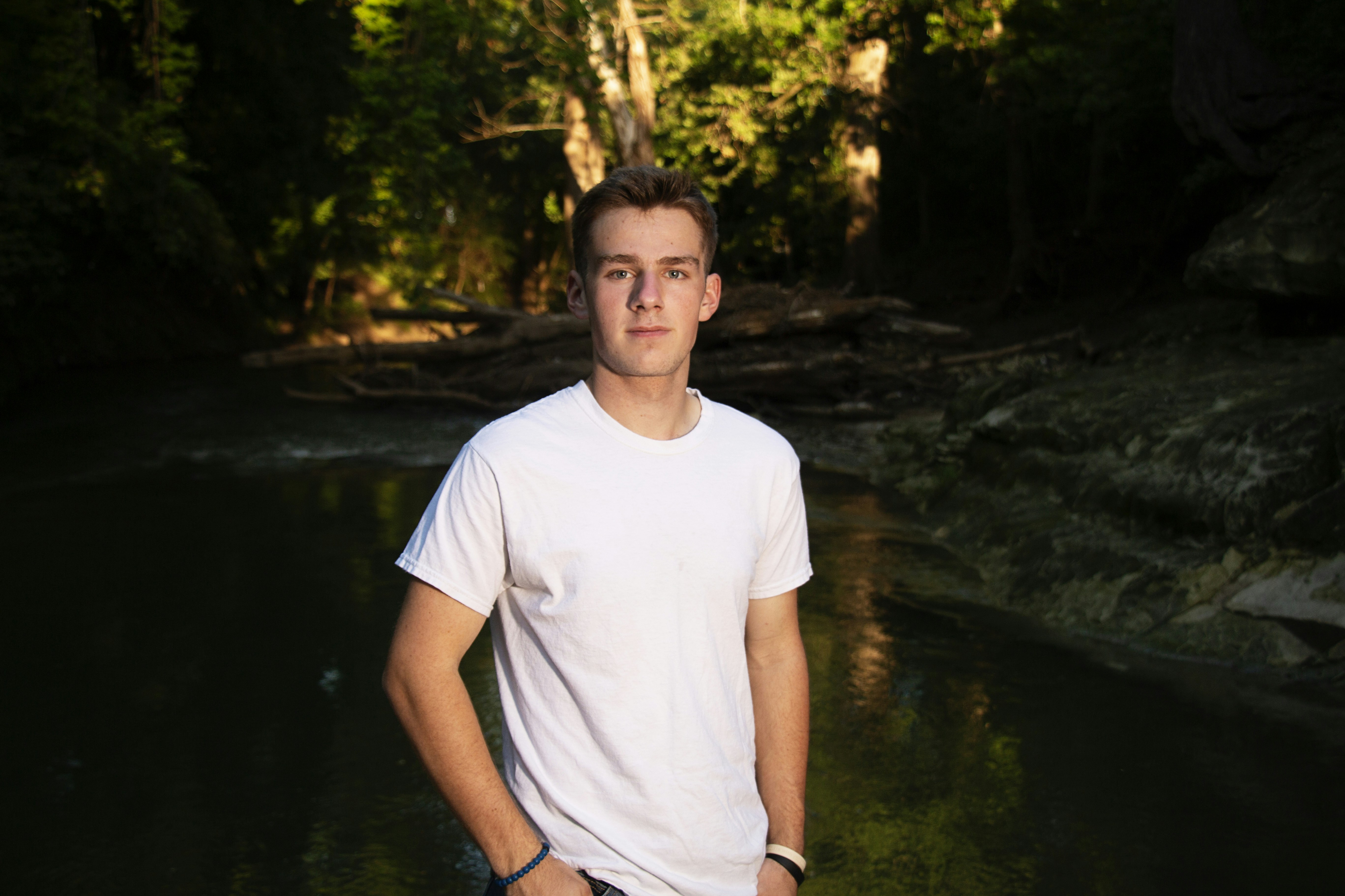 Young man standing confidently by a tranquil stream, surrounded by lush greenery. The soft light enhances the peaceful atmosphere.