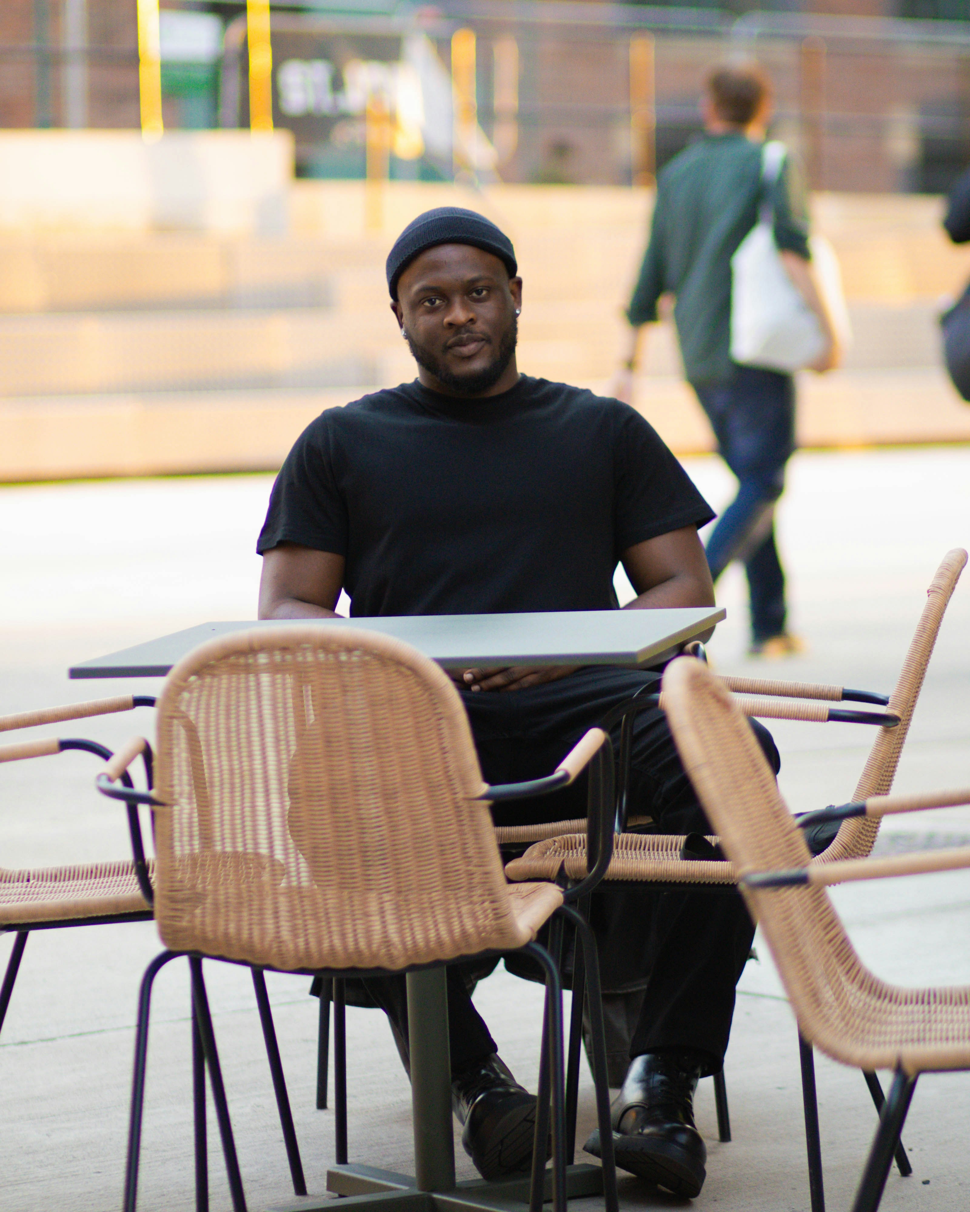 A man seated at a table surrounded by empty chairs in a lively urban setting, embodying a moment of quiet reflection amidst the bustle.