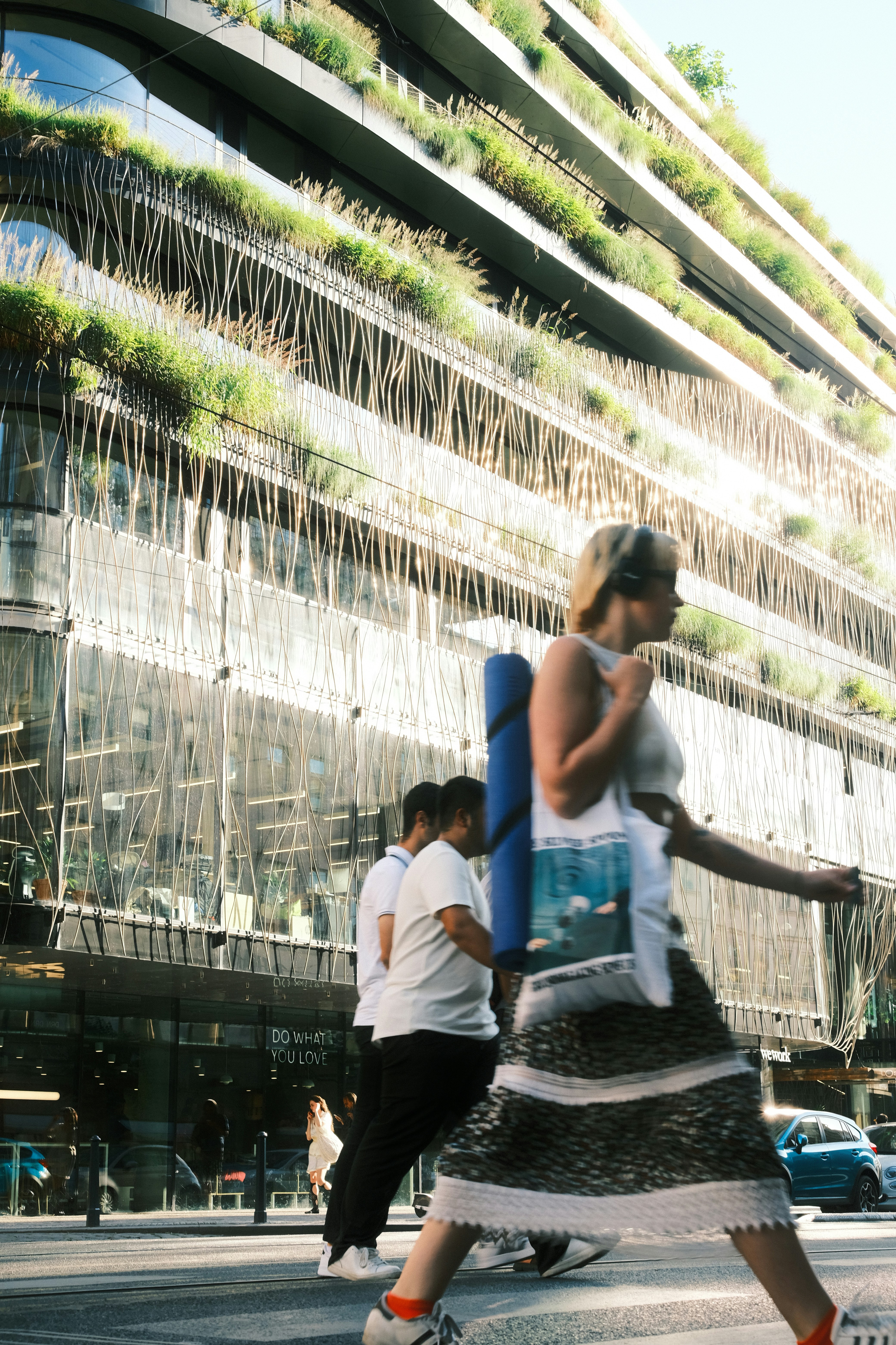Two individuals walk on a city sidewalk in front of a modern building featuring vertical gardens. One person carries a yoga mat and wears a flowing skirt. | Woman crosses street in front of modern building