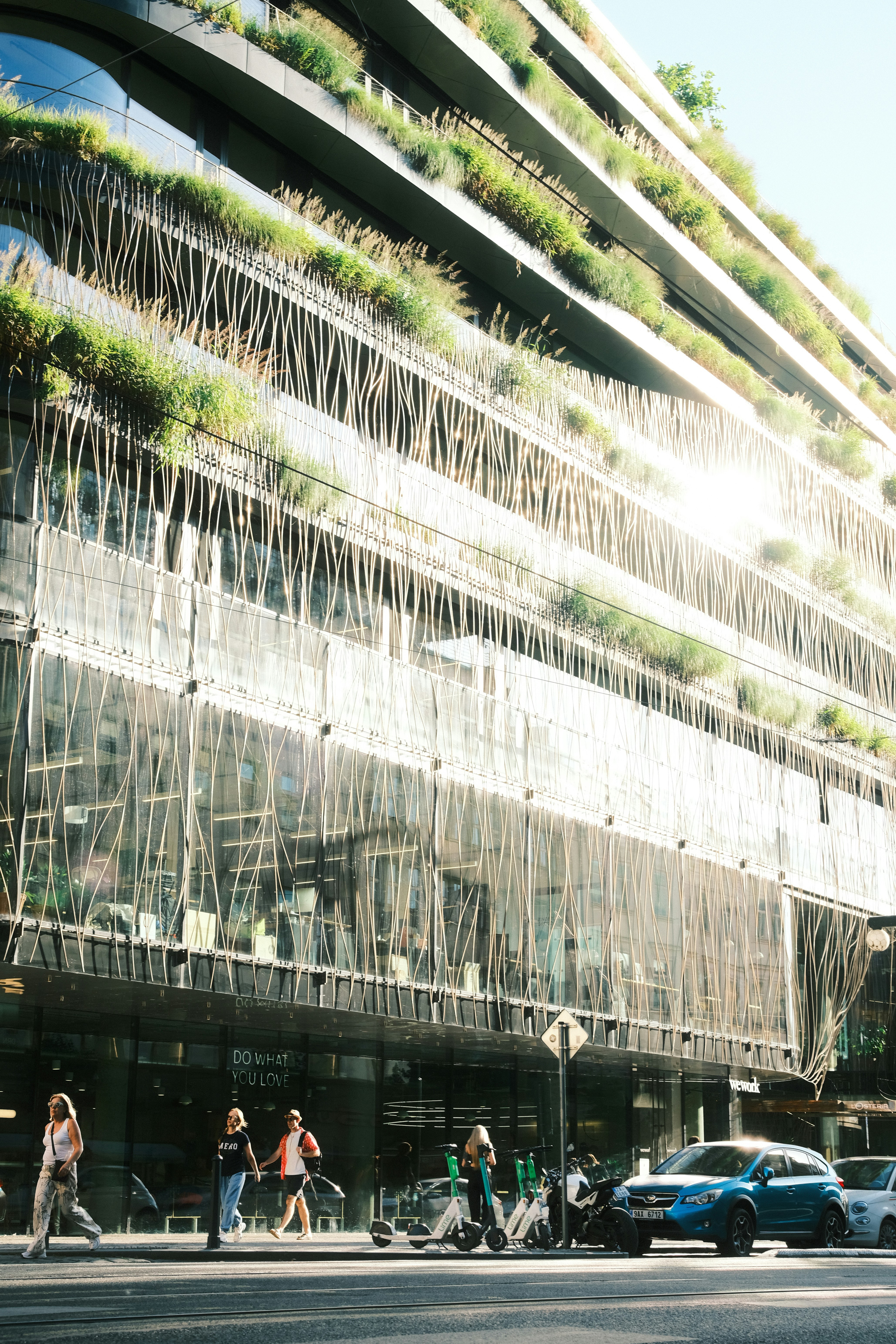 A contemporary building features a glass façade adorned with vertical greenery. Pedestrians walk along the sidewalk while cars pass by under clear blue skies. | Modern building with green terraces and glass facade