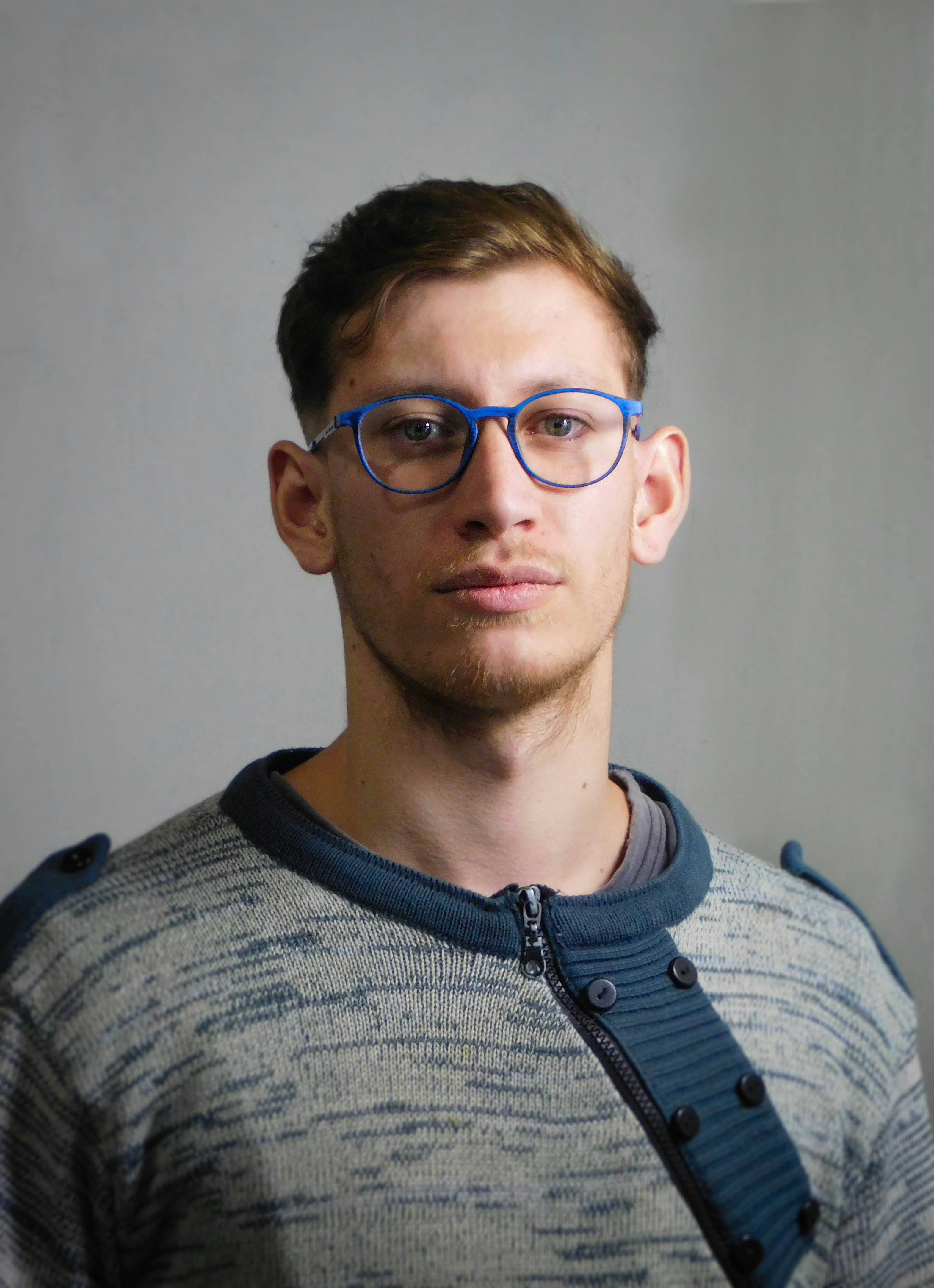 A man wearing glasses and a sweater stands facing forward.