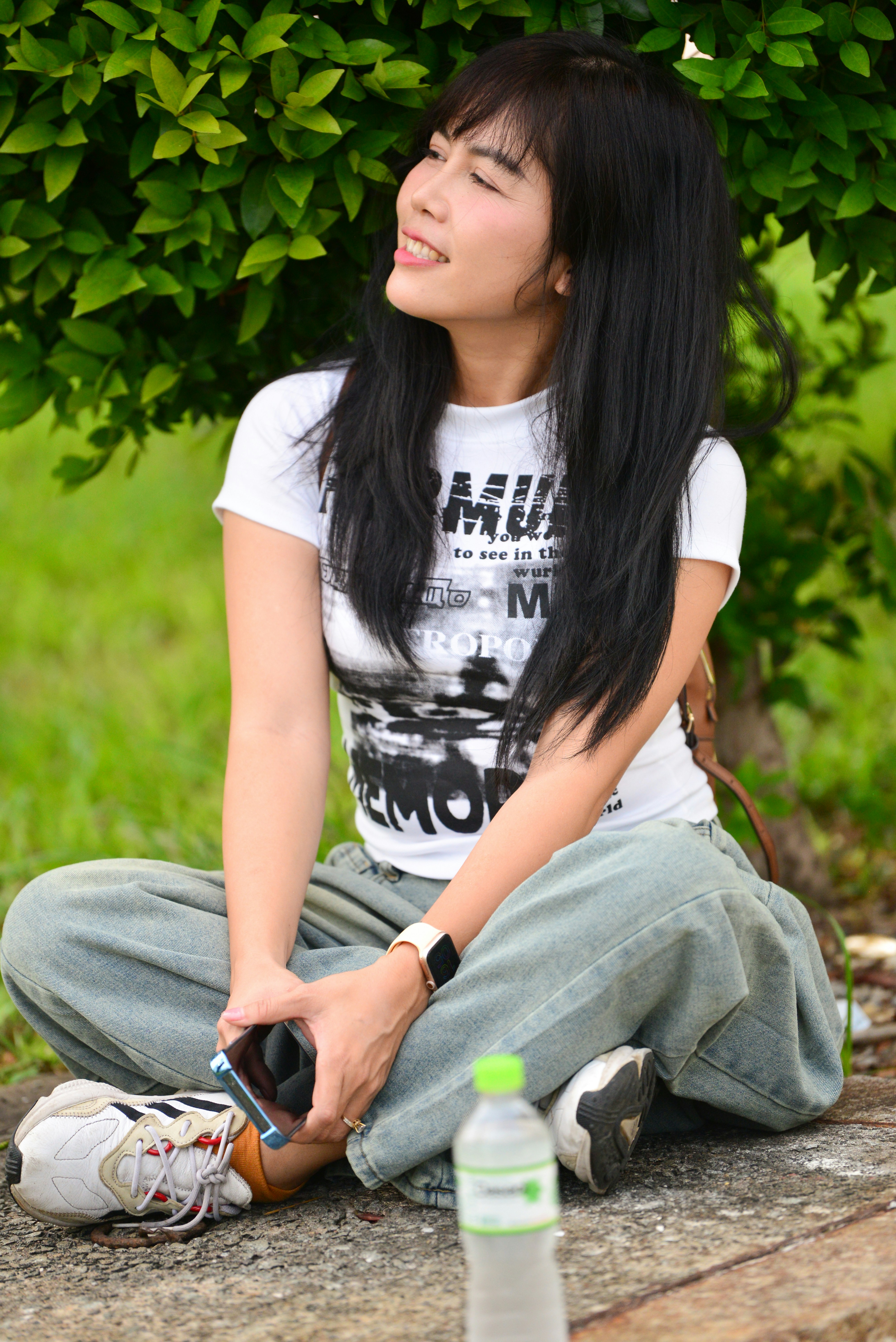 Relaxed Asian Girl Sitting on Pavement with Green Background | Young woman sitting cross-legged outdoors